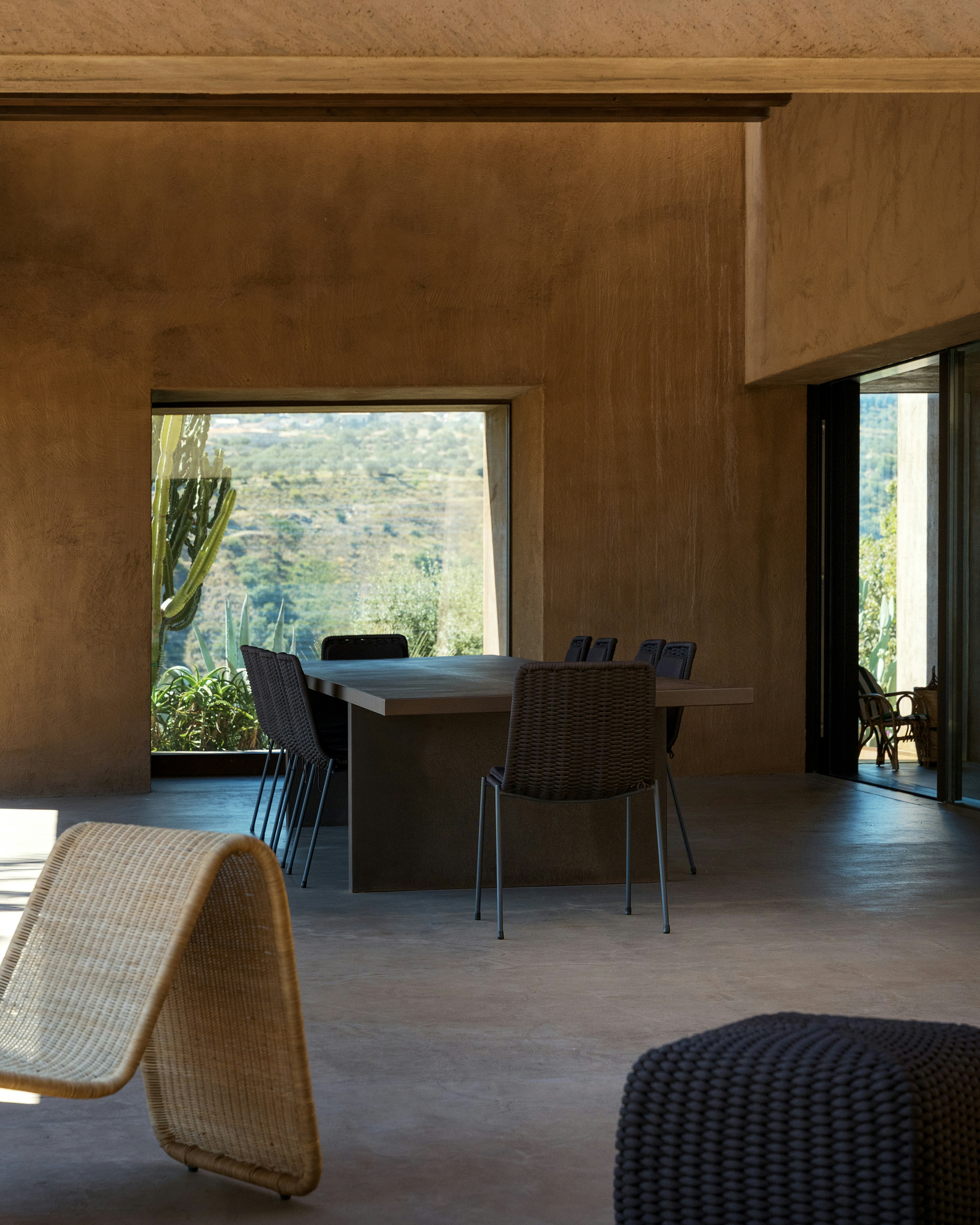 Sunlit dining area with a central wooden table, wicker chairs, and large windows revealing greenery outside. A calm, architectural interior emphasizing natural materials.