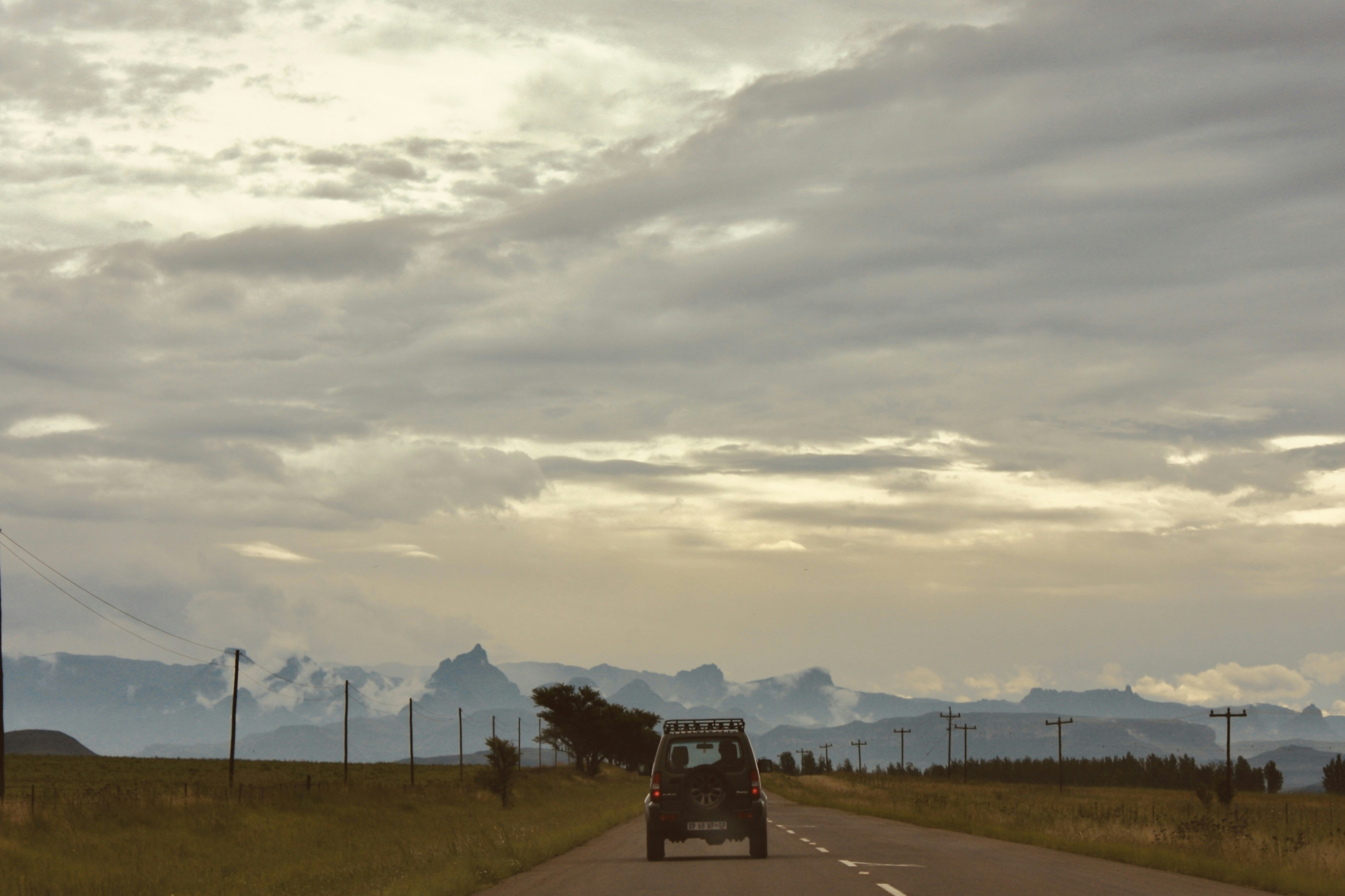 Car travels a straight rural road under a wide, cloudy sky; the scene emphasizes open space and distant mountains.