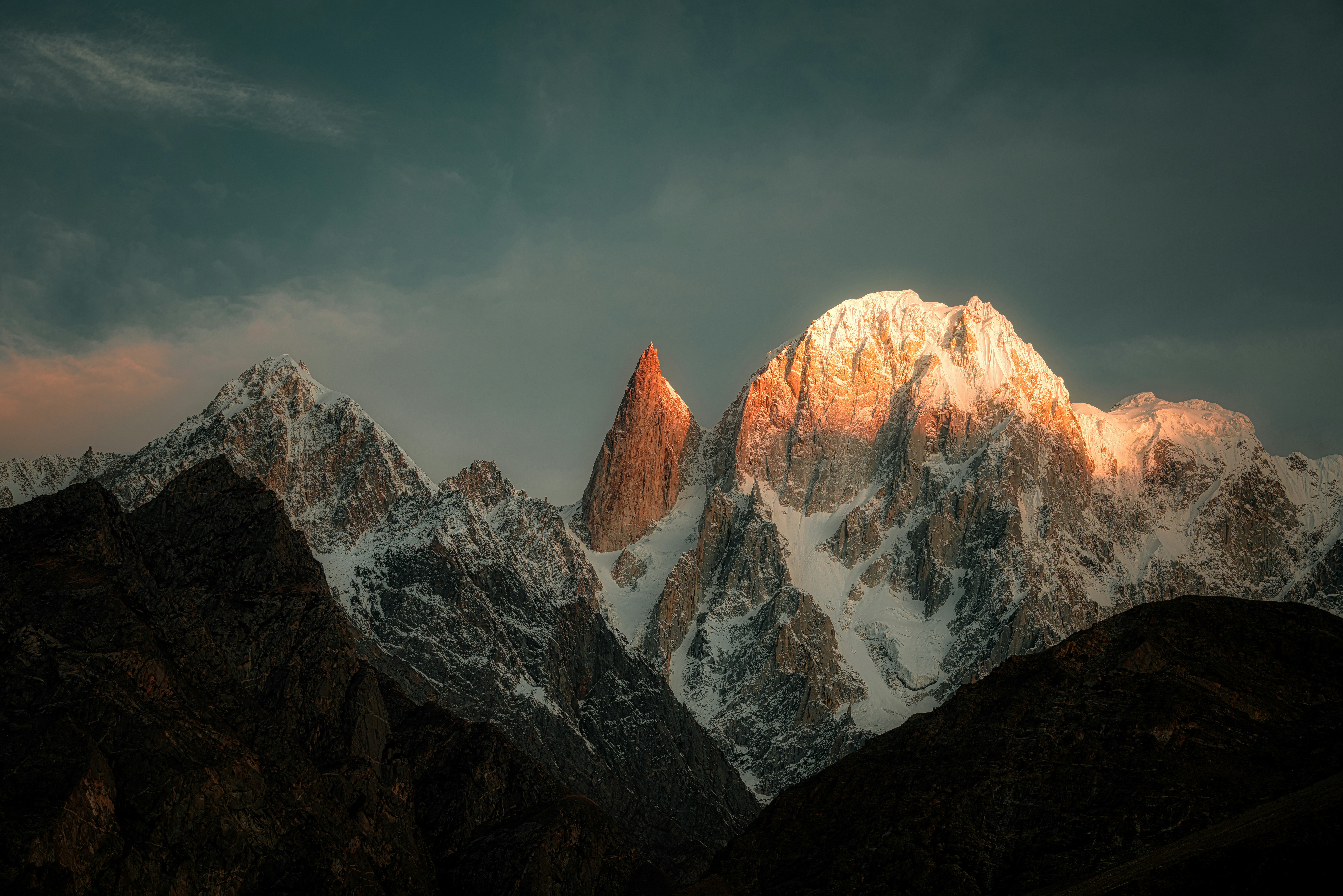 Snow-capped peaks bathed in warm dawn light rise above dark foothills; a dramatic alpine scene highlighting rugged textures and subtle color gradient.