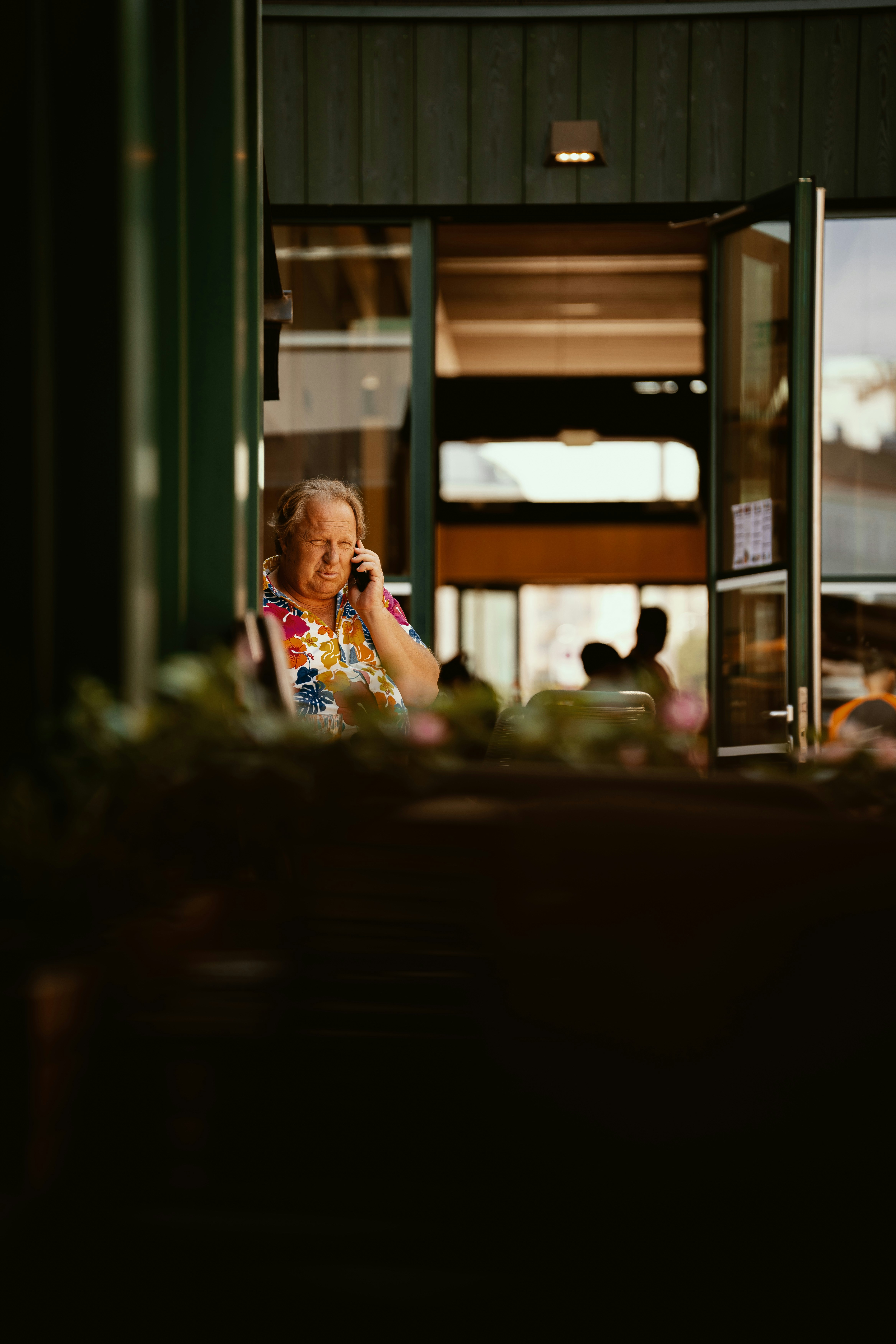 A warm, candid portrait of an elderly woman in a colorful shirt talking on a mobile phone at a cafe doorway; the foreground is blurred with a shallow depth of field, emphasizing her relaxed expression and the interior light behind.