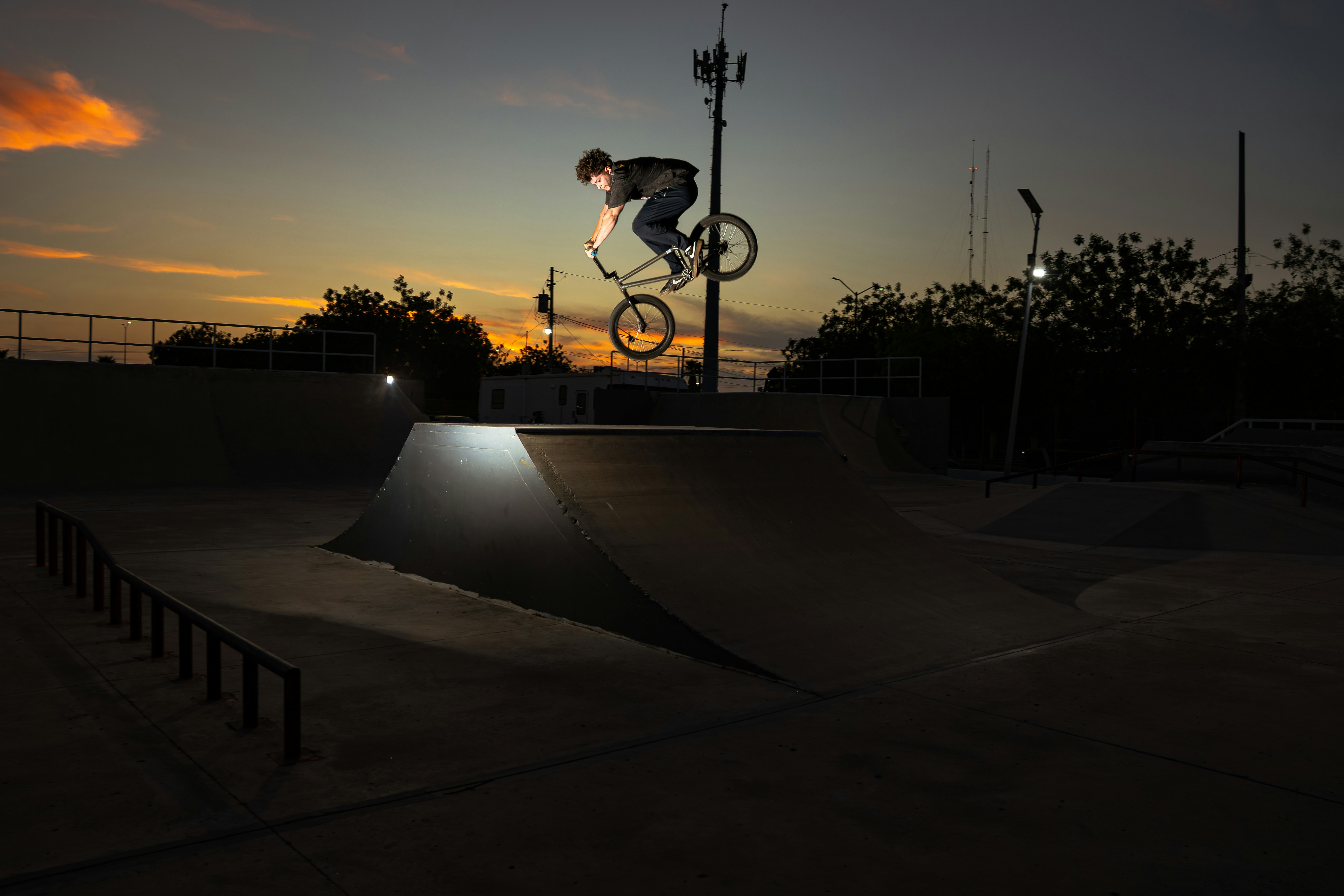 A BMX rider soars off a ramp at dusk in a skatepark, silhouette against the fading sun and artificial lights; the scene hints at a deliberate, action-filled moment in a controlled environment.
