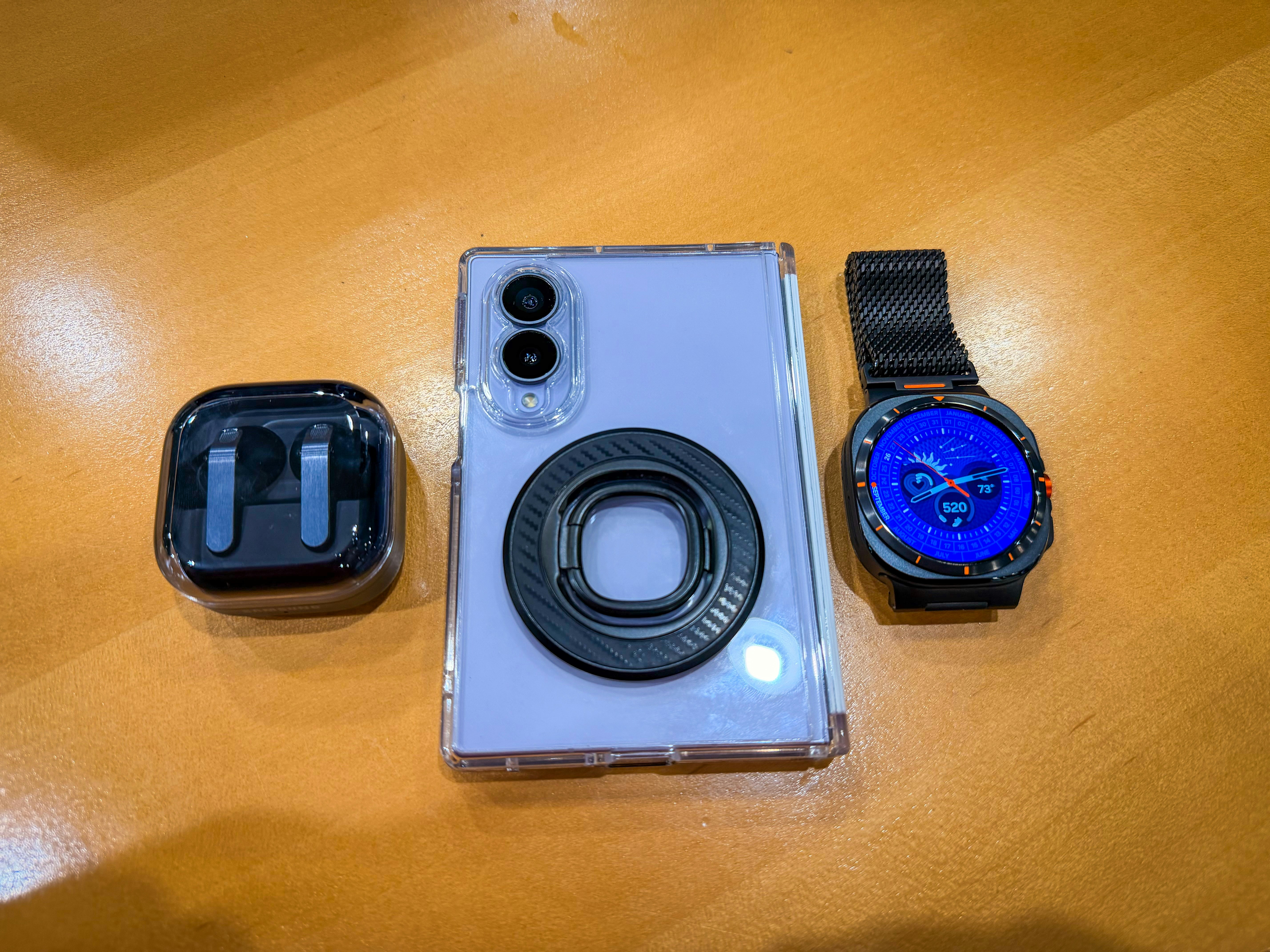 Three everyday tech items arranged on a wooden table featuring a wireless earbuds case, a clear phone case around a device, and a smartwatch with a blue-accented dial.