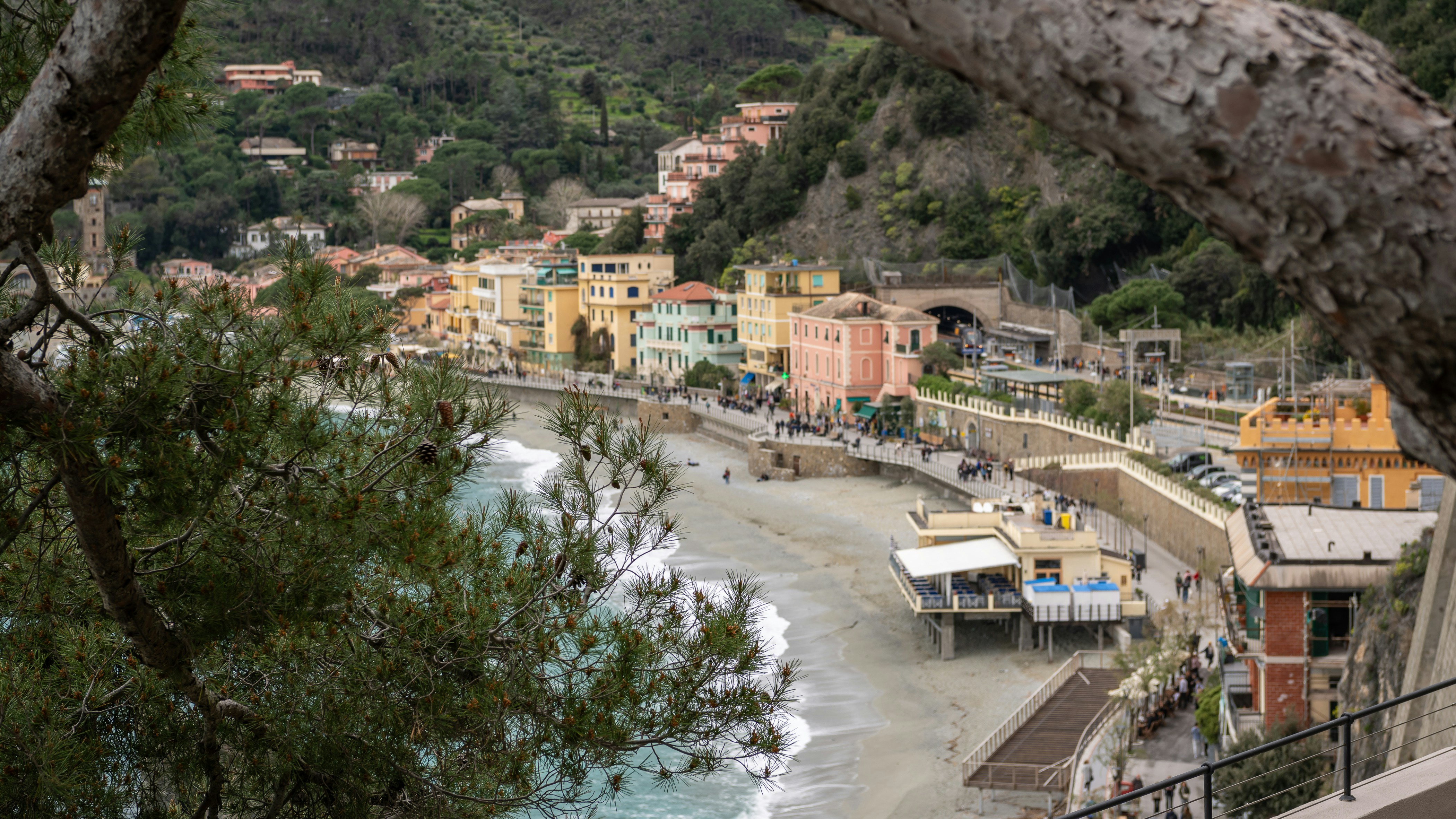 Colorful coastal village along a curving beach with pastel buildings and a boardwalk, framed by hillside trees and distant boats.
