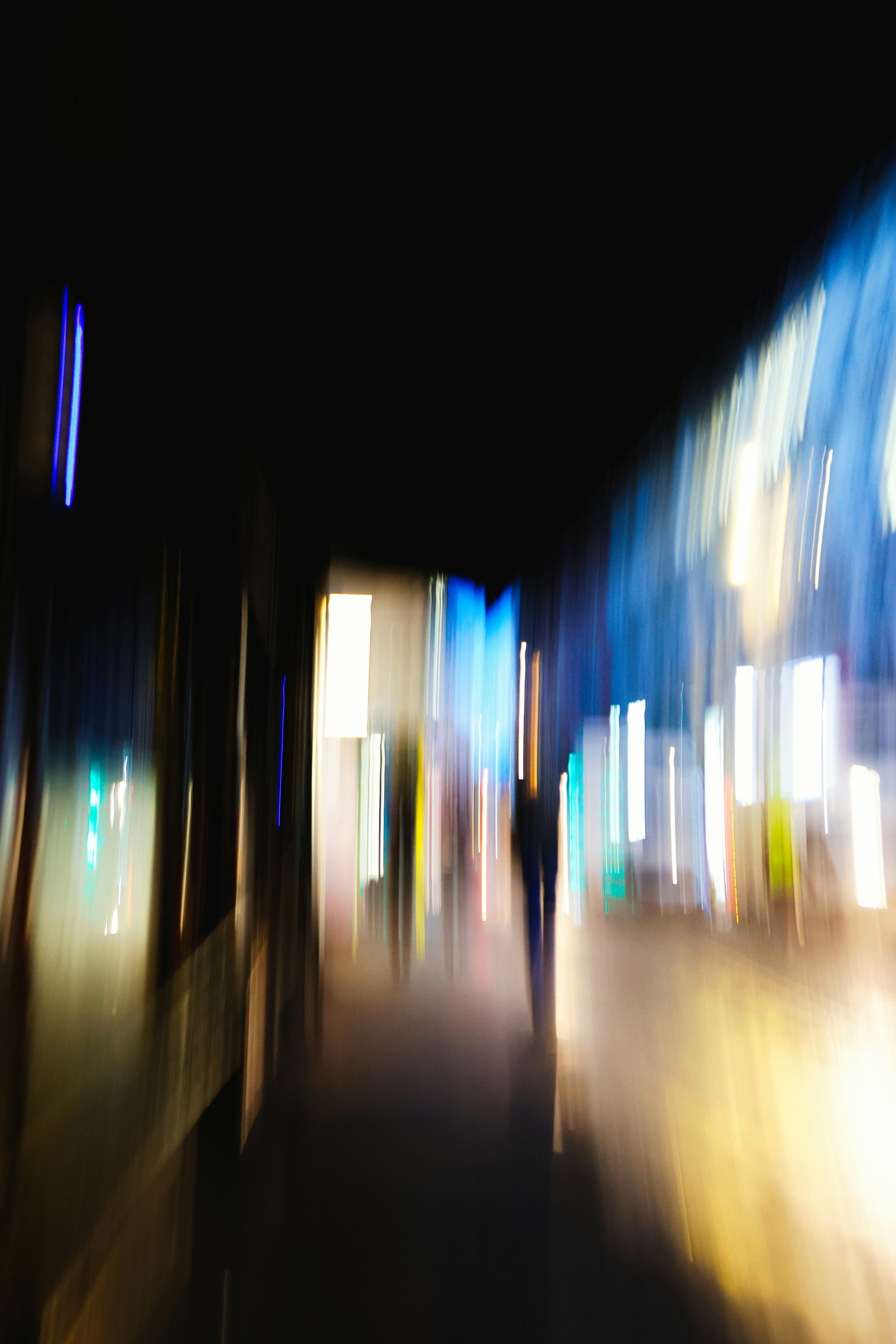 Urban night scene with motion blur and streaked lights, suggesting movement and energy in a city street. The blurred figure in the center adds a human scale to the urban chaos.