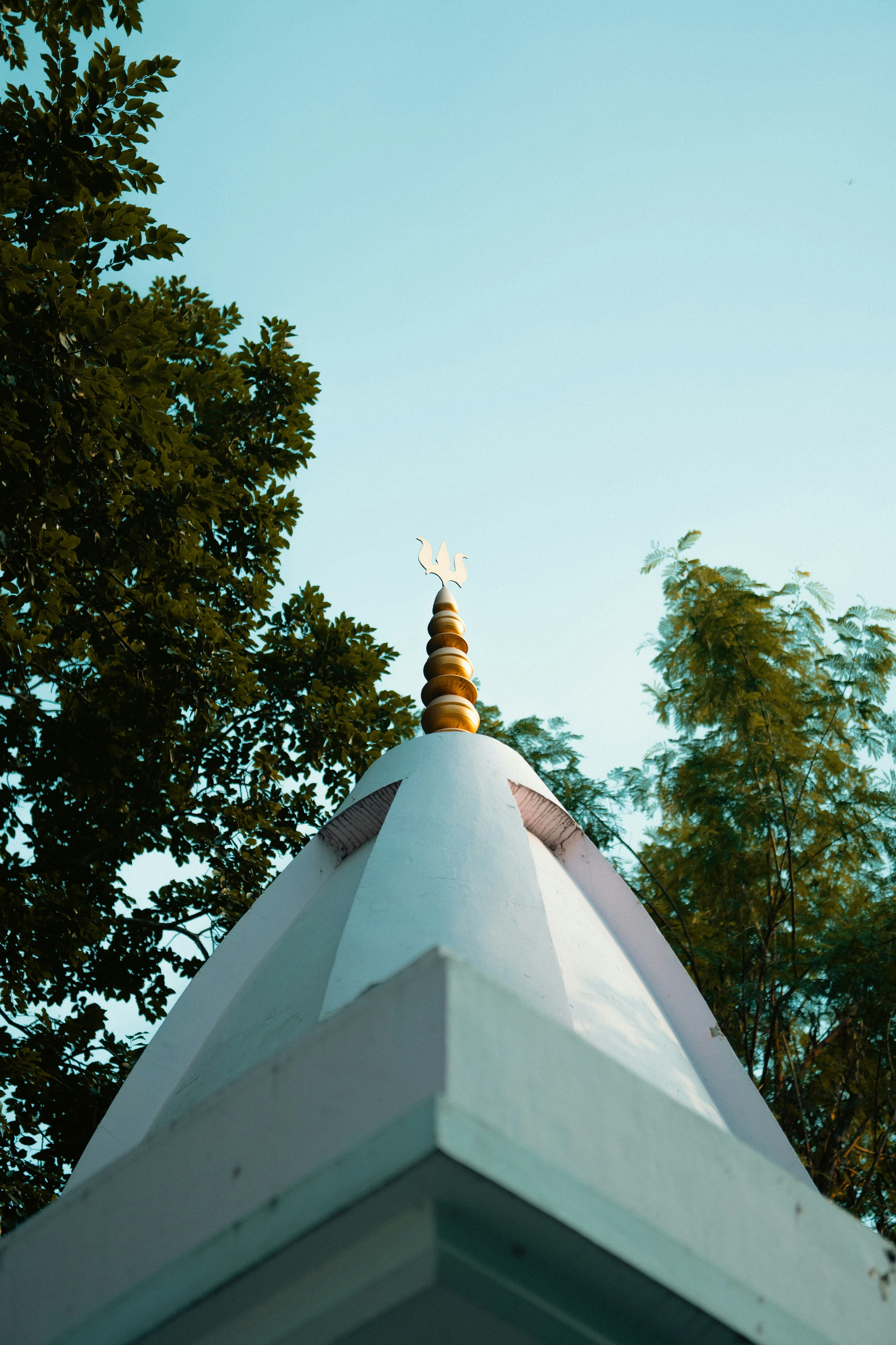 White temple spire with gold finial rising between green trees against a pale blue sky.