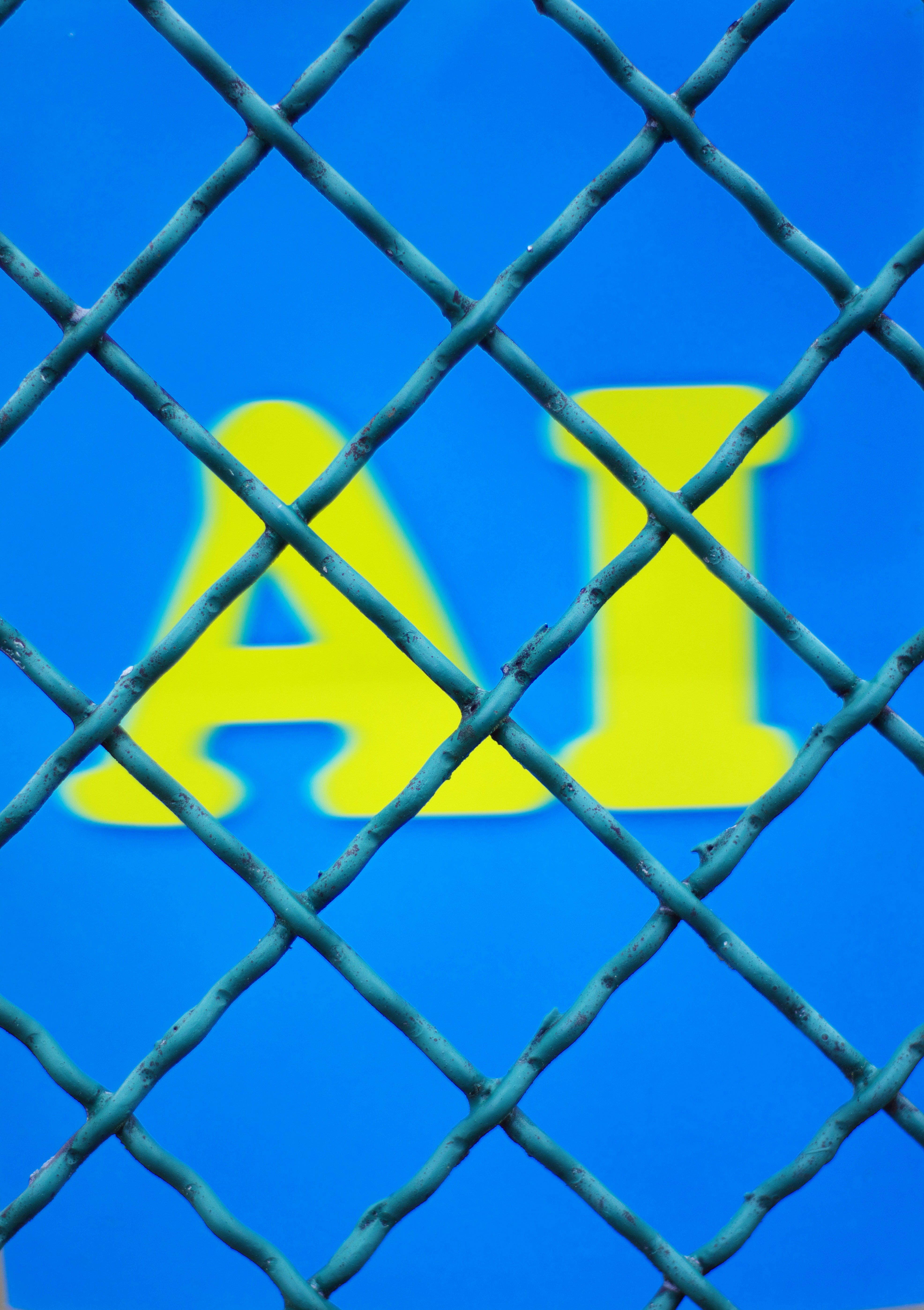 Blue backdrop with bold yellow letters seen through a diamond-pattern chain-link fence; the subject is the blurred signage behind the fence, emphasizing texture and color contrast.
