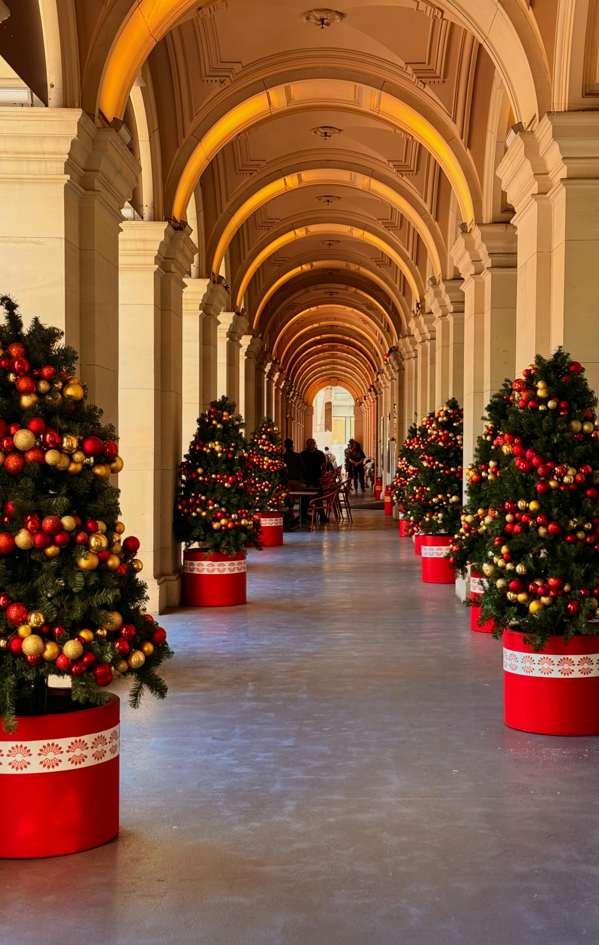 Holiday corridor lined with decorated Christmas trees under an arched ceiling, inviting center path leading to a distant light; festive scene with warm lighting.