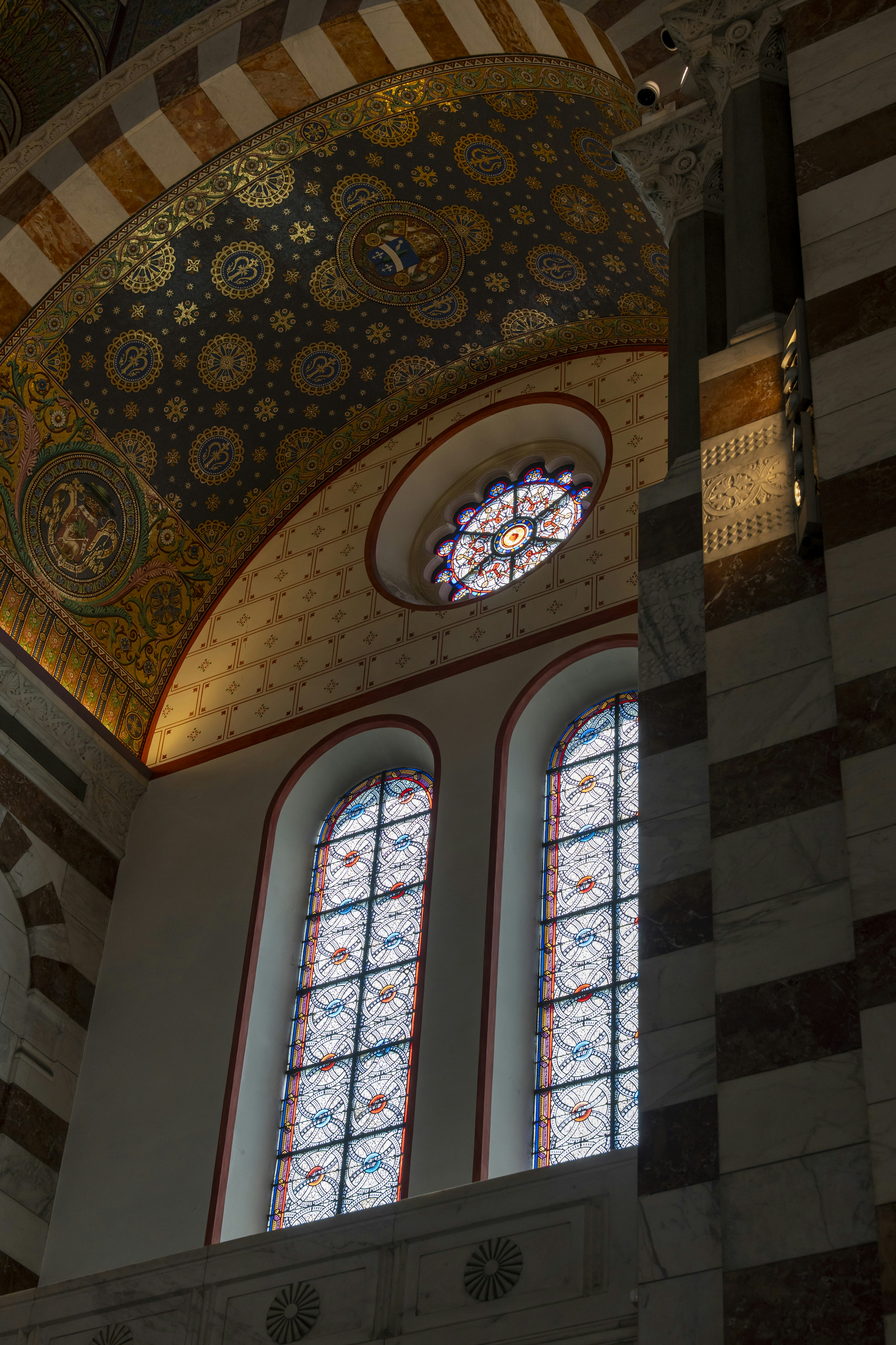 Sunlit stained glass windows rise beside marble arches beneath ornate patterned ceilings, highlighting ecclesiastical architecture.