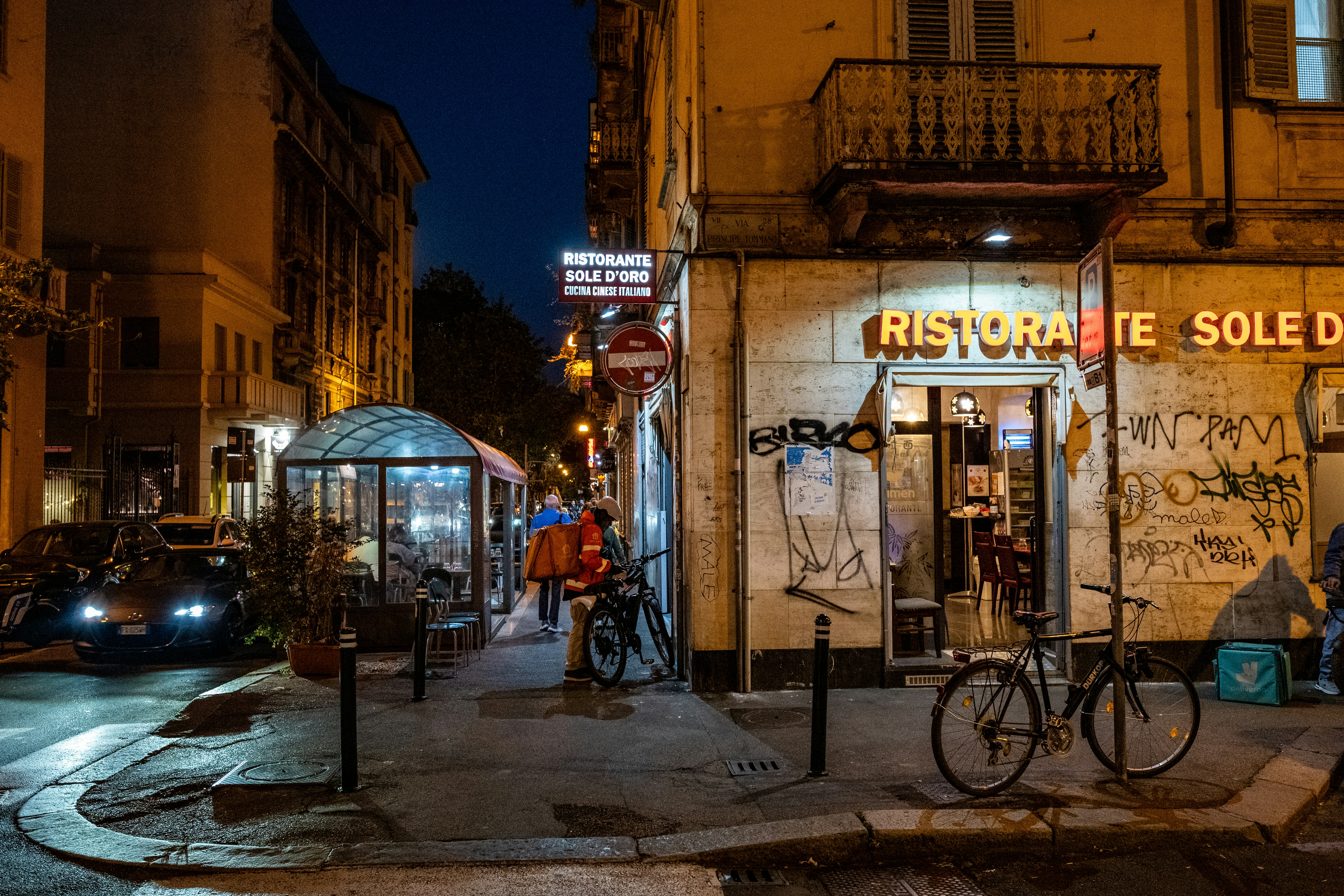 Night street scene with a lit restaurant façade, outdoor seating, and bicycles; warm neon signage contrasts with blue ambient light.