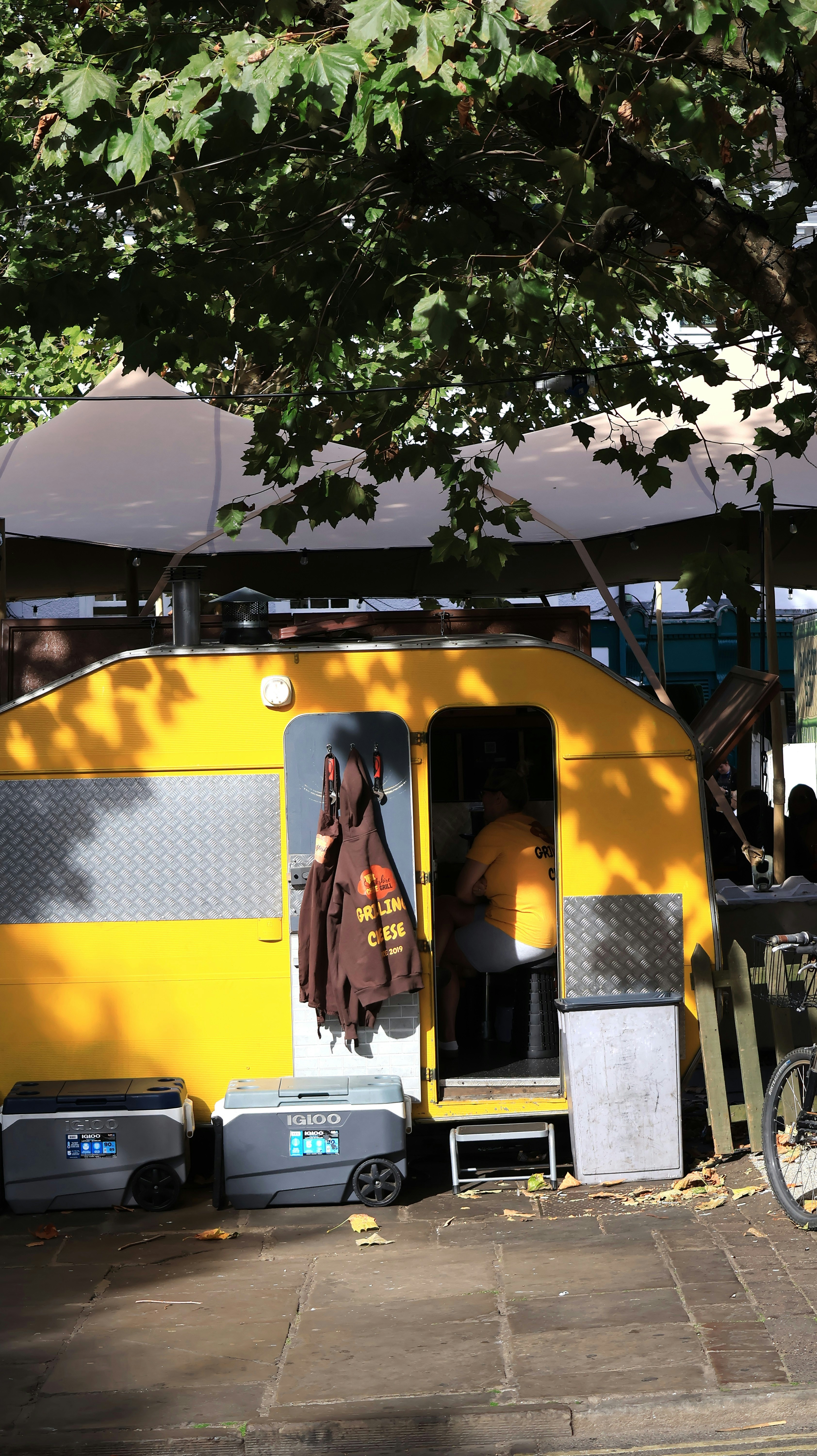 Bright yellow caravan goods trailer under leafy canopy with a person in a yellow shirt seated at the door; tools and coolers sit on the ground nearby. A jacket hangs on the door and a bicycle is partially visible.