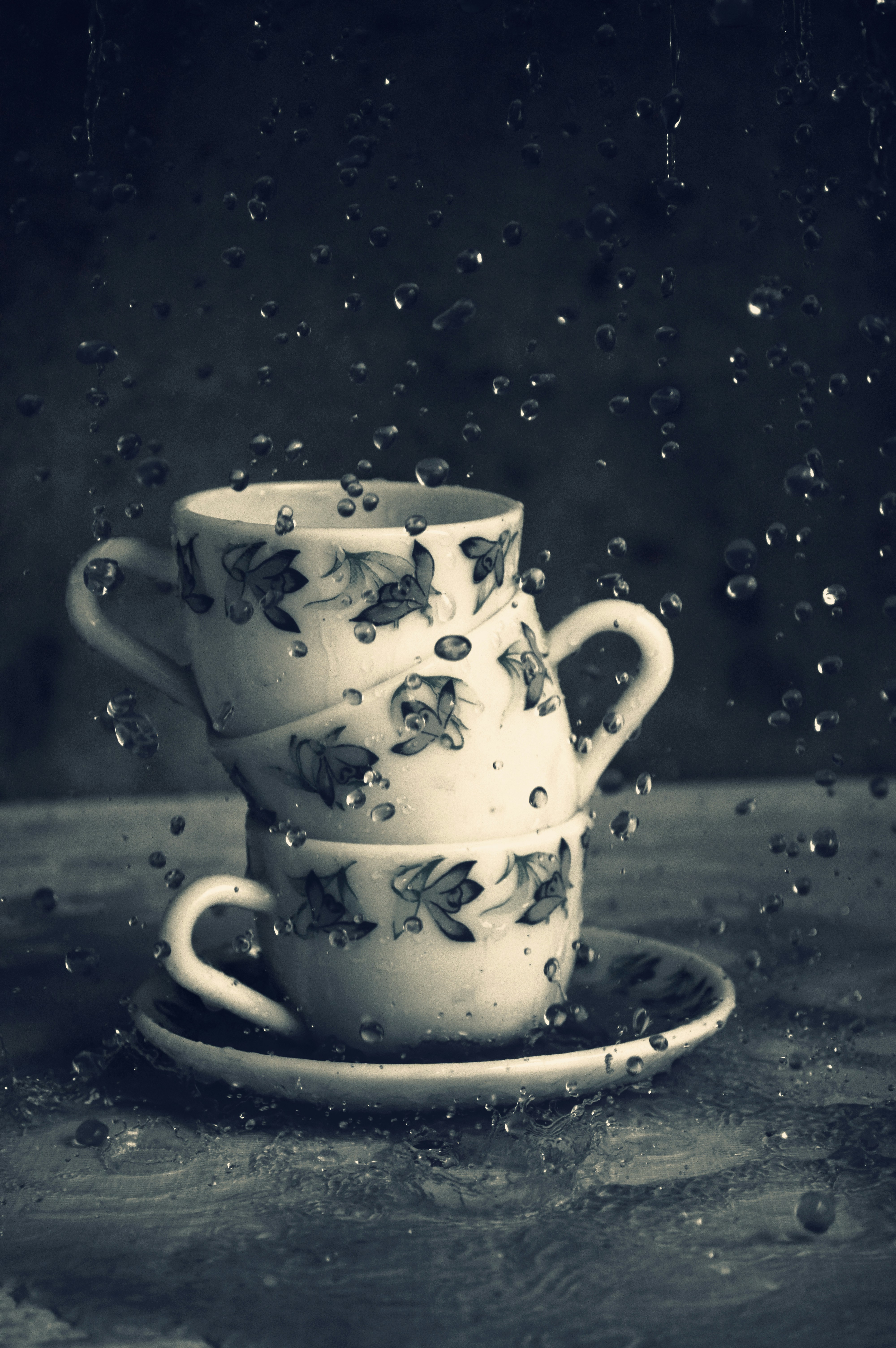 Stacked floral teacups sit on a saucer as rain-like droplets cascade across the scene, creating a moody still-life with a soft, vintage tone.