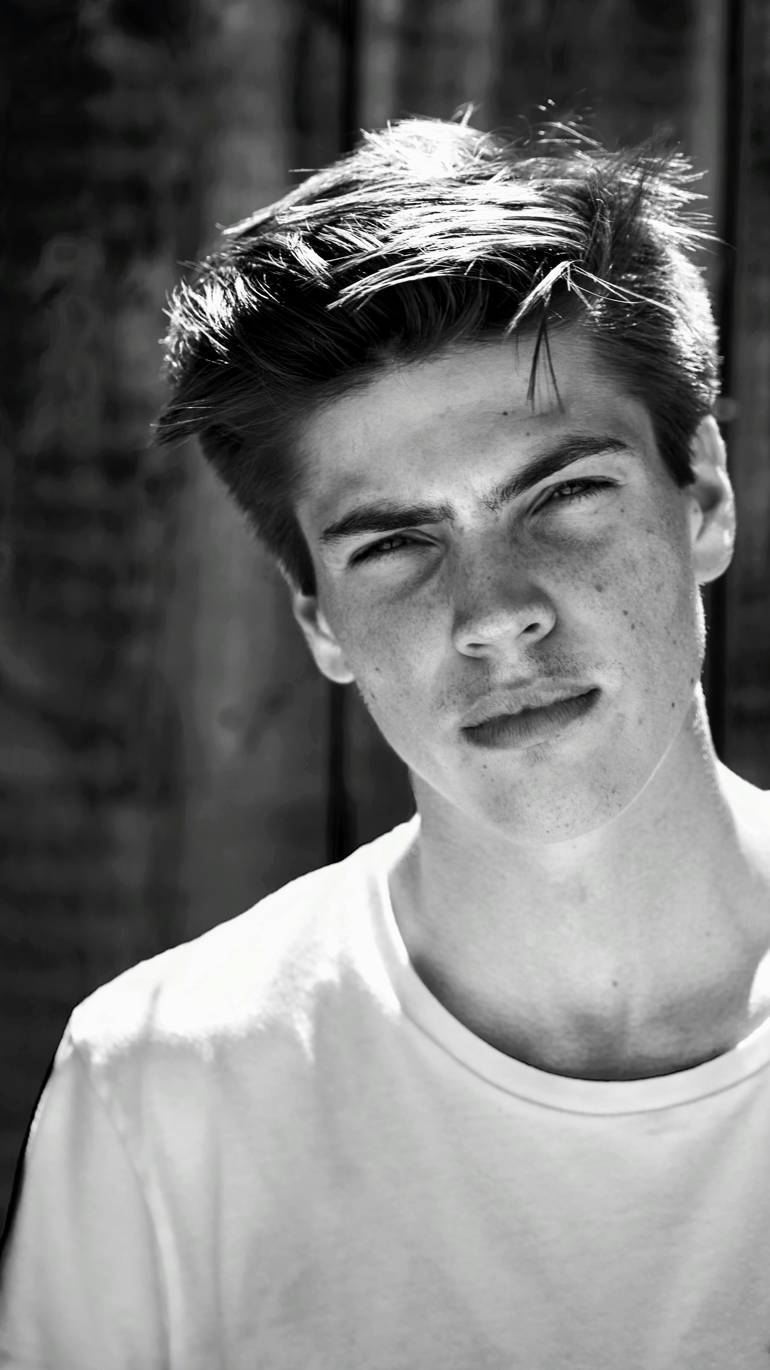 Monochrome portrait of a young man with a focused gaze, sunlight catching wind-swept hair, against a blurred wooden background. This image emphasizes facial features and expression.