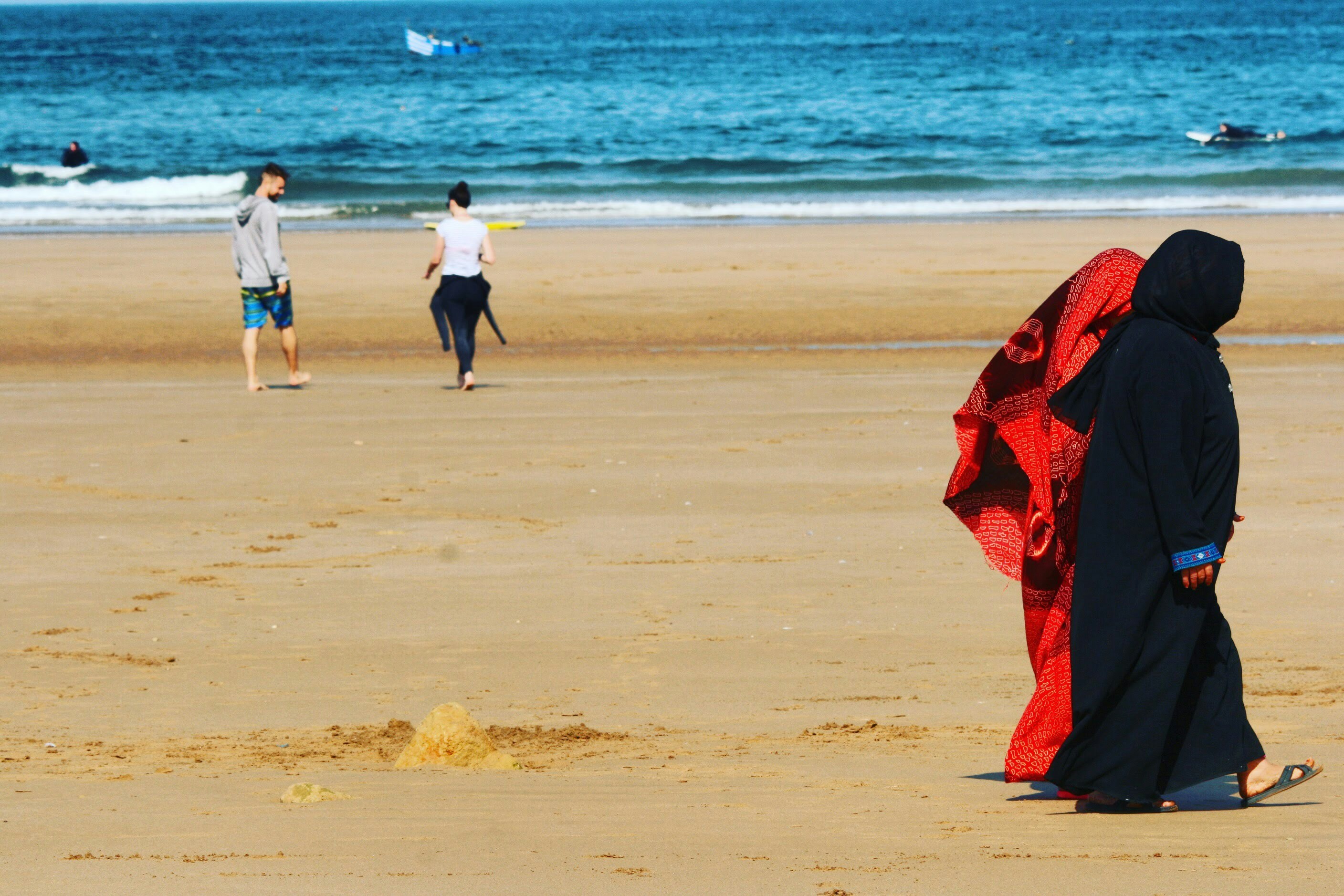 A lone veiled figure in dark robes with a red scarf walks along a sunlit beach, while two people wander near the shoreline and the sea stretches into the horizon.
