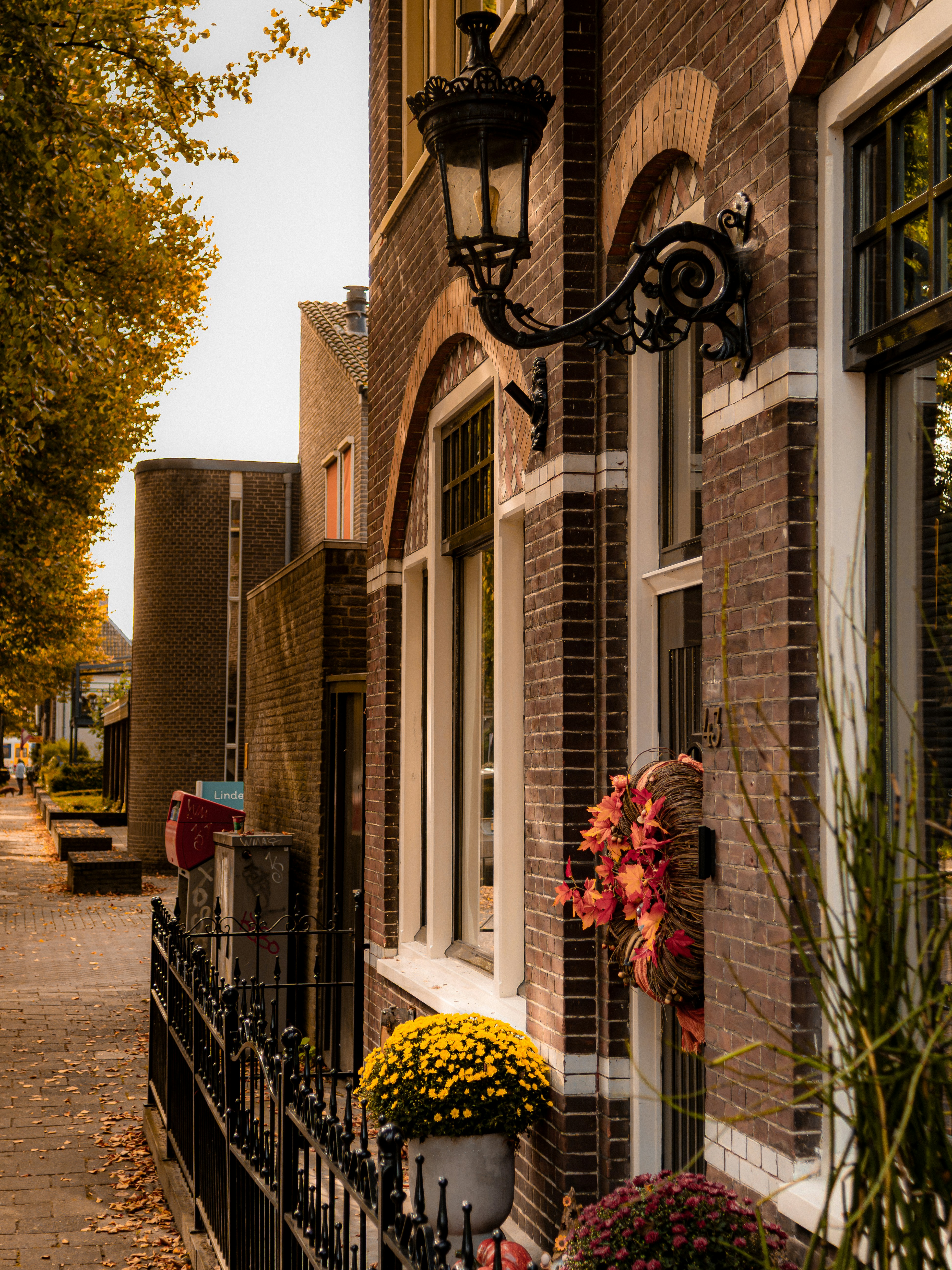 A close-up perspective of a traditional brick townhouse facade in the Netherlands during the fall season. The scene features an ornate, vintage black iron street lantern mounted on the wall, a colorful autumn wreath hanging on the front door, and a vibrant pot of yellow chrysanthemums by a classic wrought iron fence.