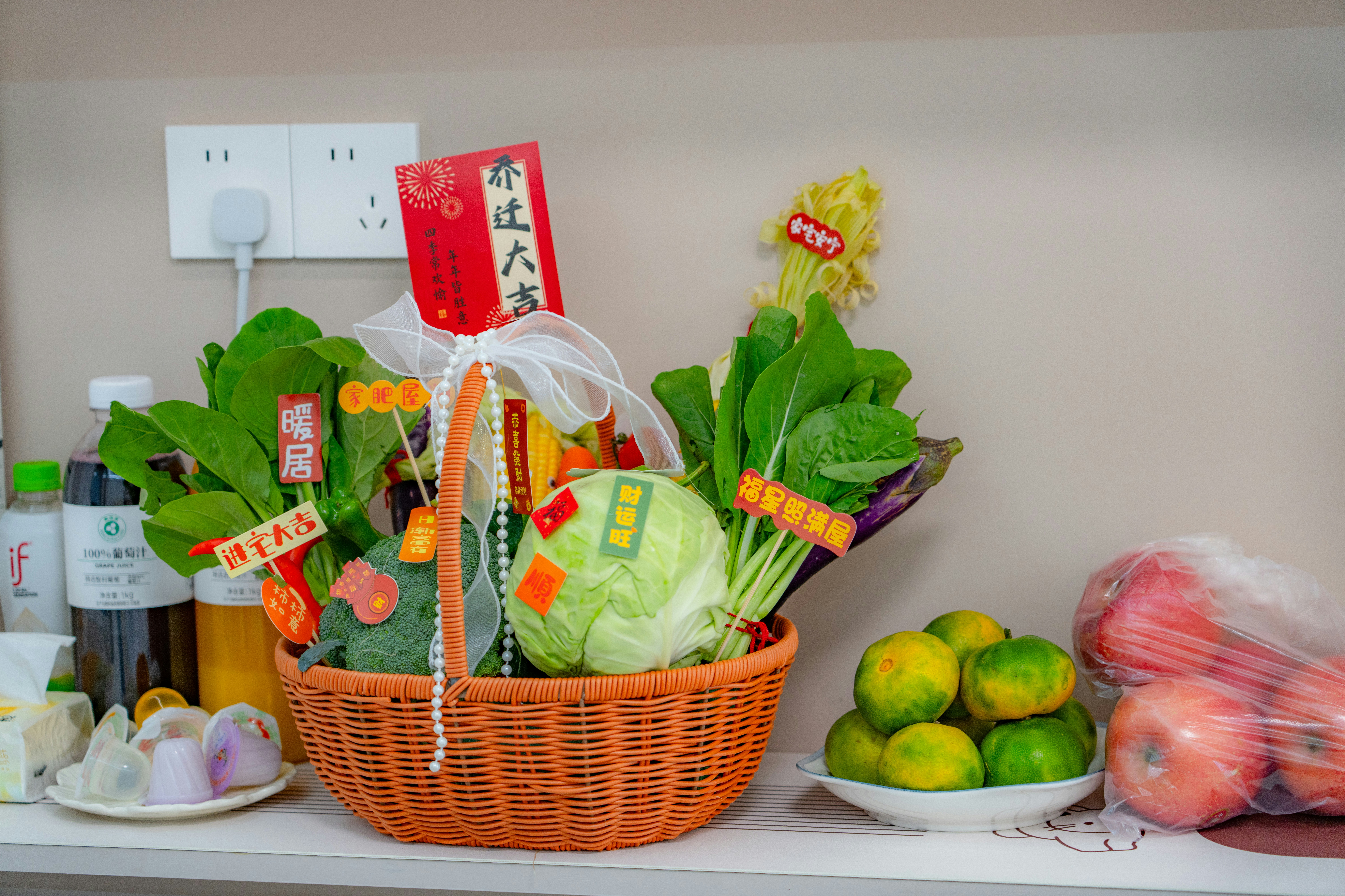 Assorted vegetables and fruits arranged in a wicker basket with red tags and leafy greens on a kitchen counter, alongside additional produce in the background.