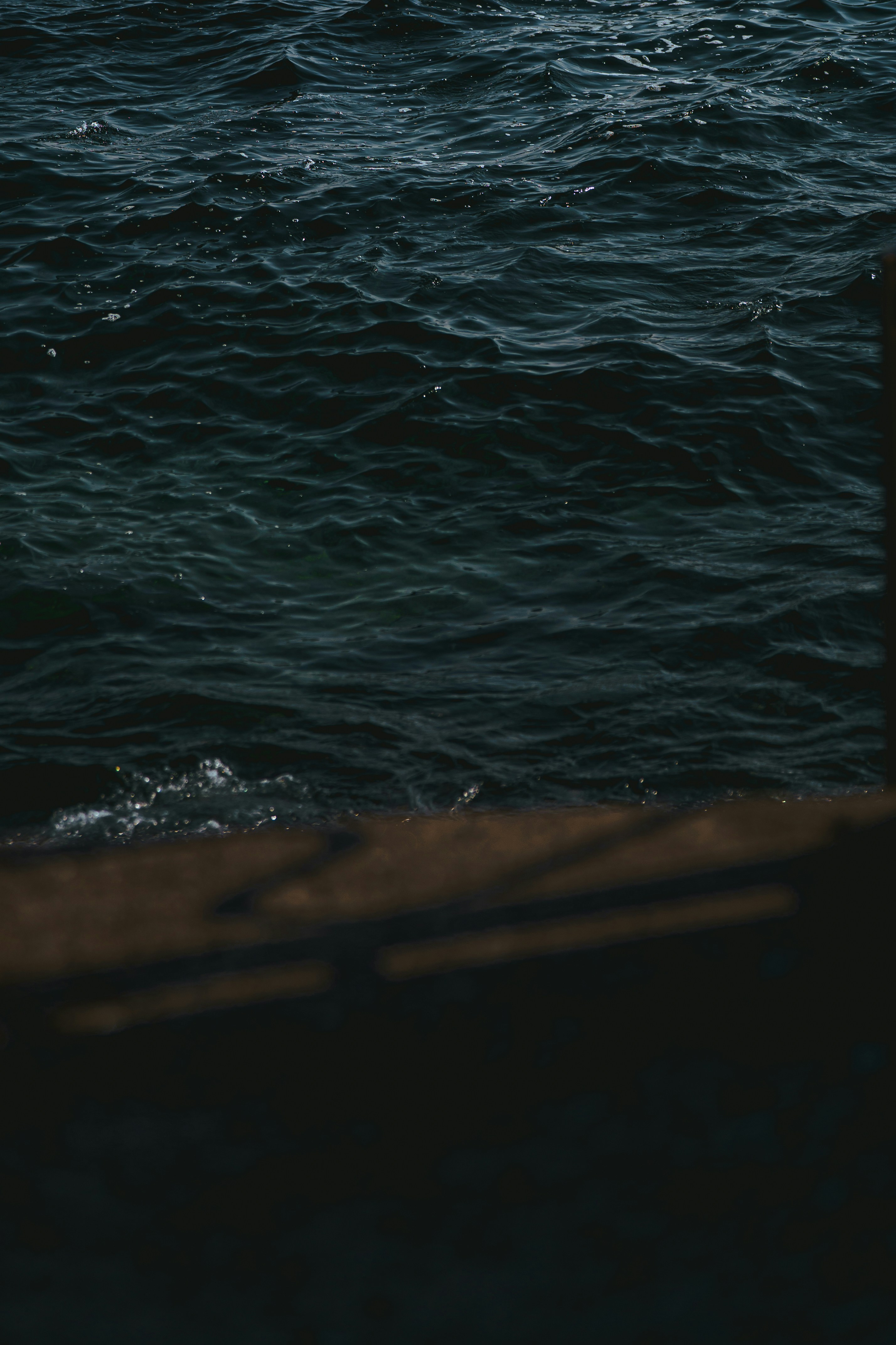 Dark, teal sea ripples along a concrete edge; the image emphasizes the boundary between water and shore with subtle motion and low light. This is a photographic capture, not a drawing or painting.