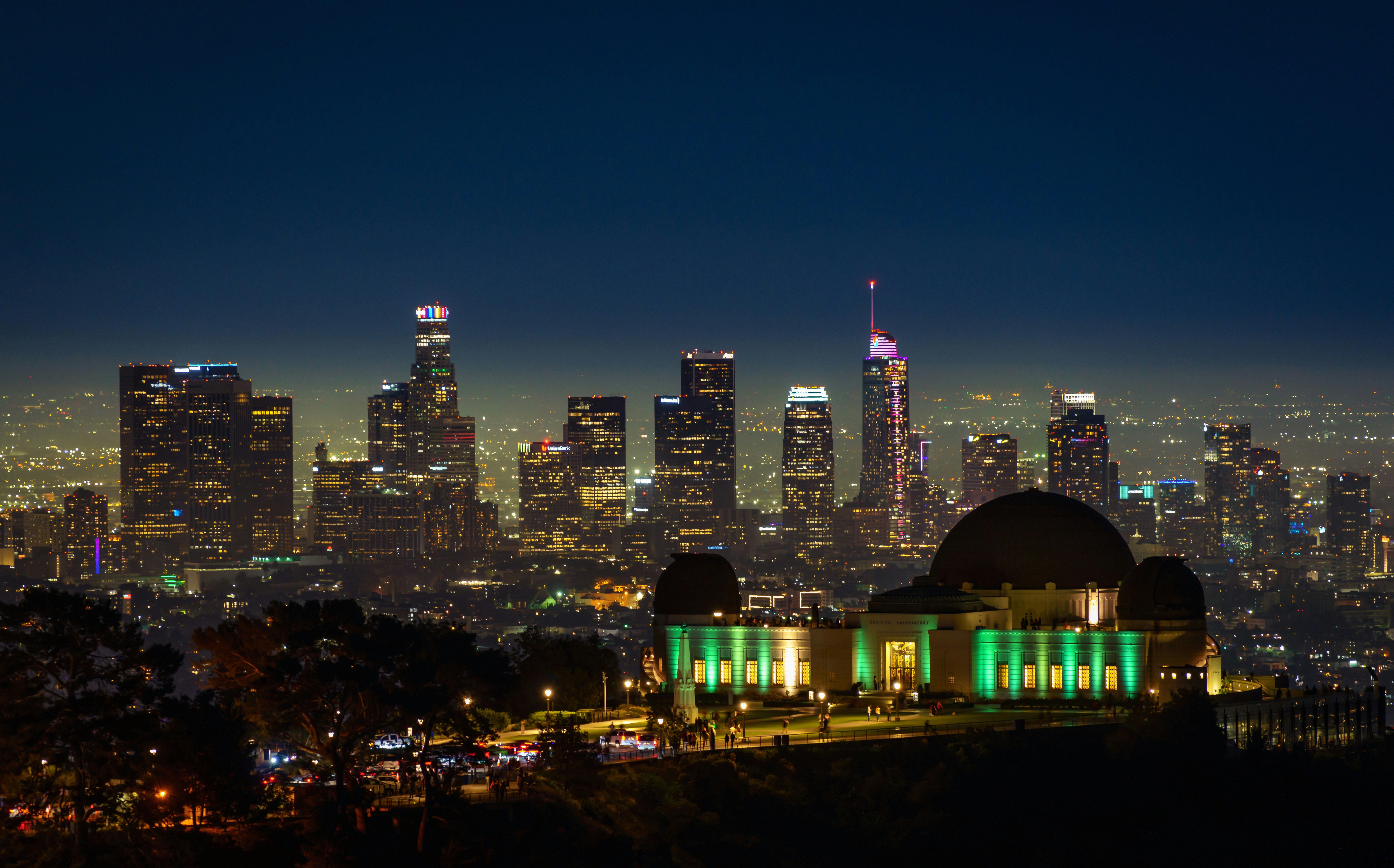 Night view of a city skyline from an elevated observatory with green-lit dome structures in the foreground.