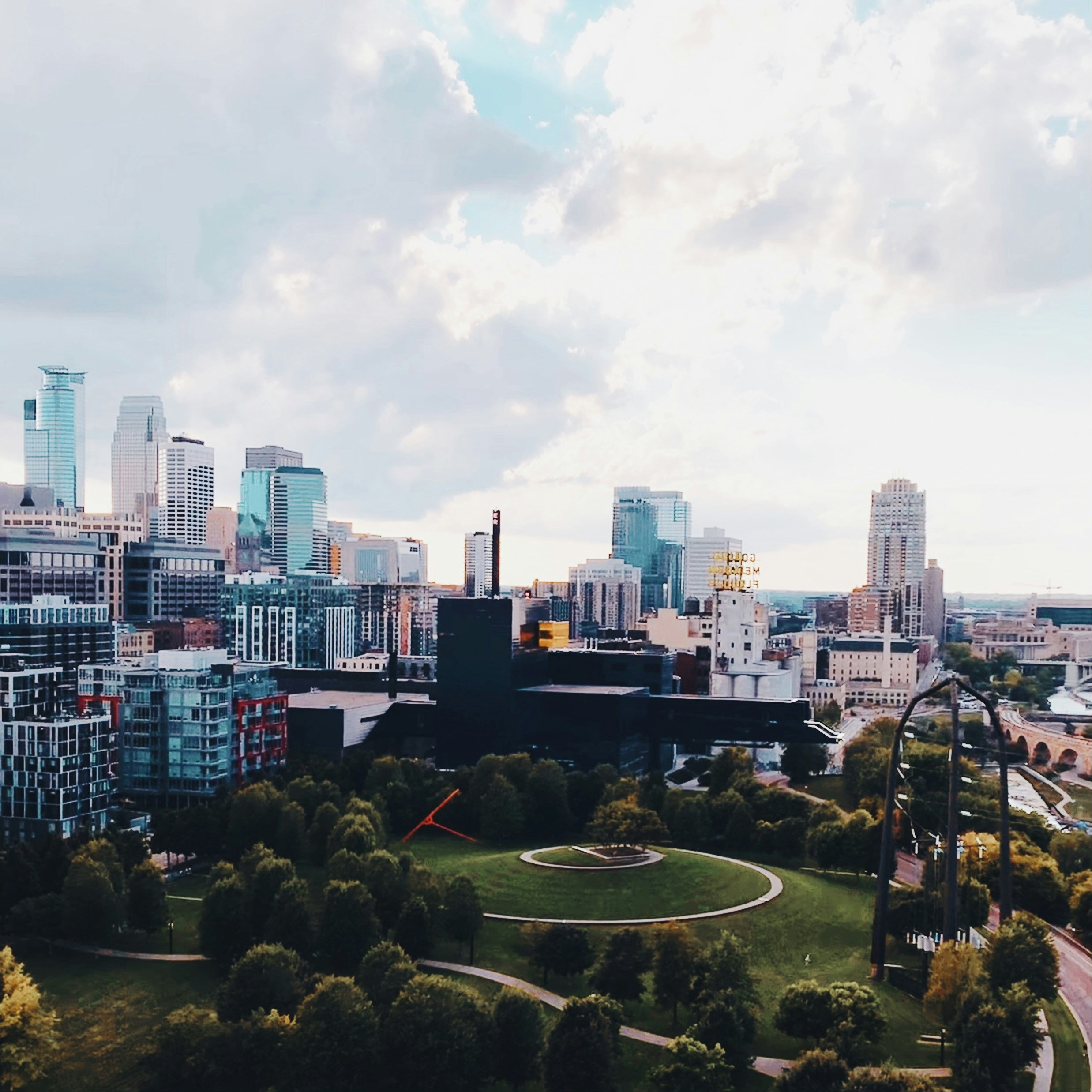 A sprawling urban skyline rises beyond a looping circular park with paths and trees, captured in daylight.