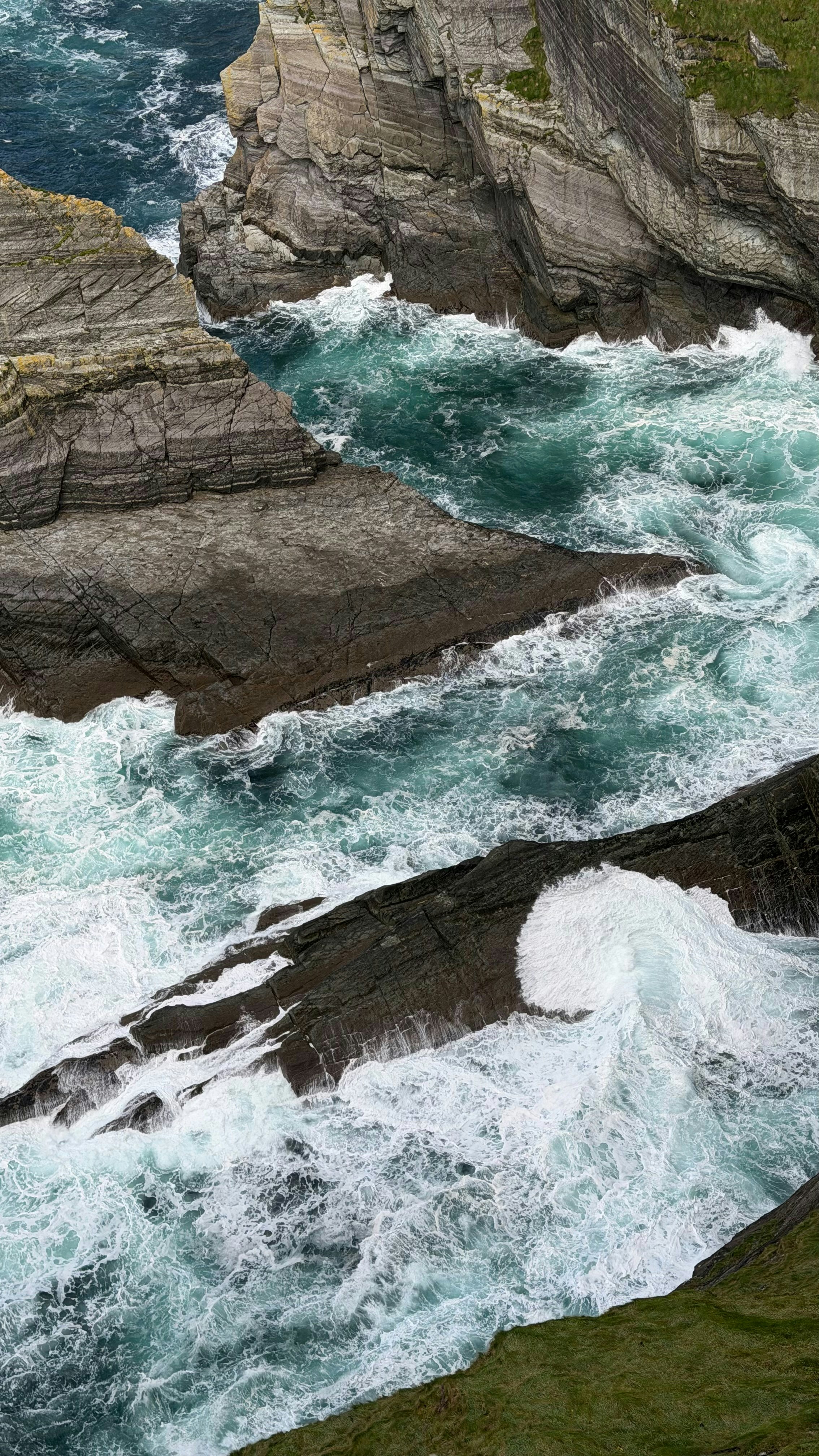 Cascading turquoise sea cuts through jagged coastal rock faces, revealing a dramatic shoreline and foamy white waves. Aerial perspective highlights the rugged geology and dynamic water motion.
