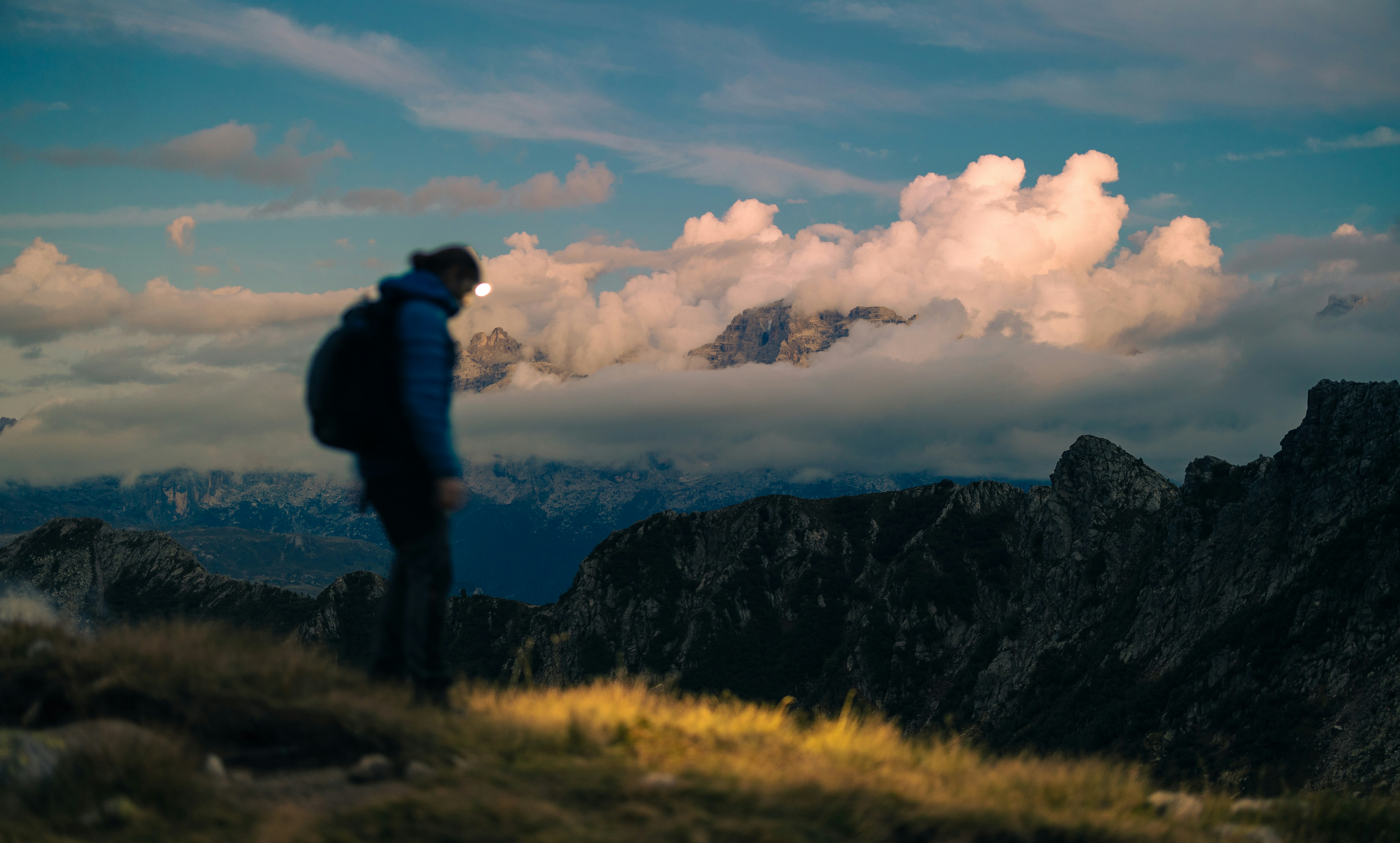 A lone hiker with a backpack traverses a misty alpine ridge as clouds drift over jagged peaks, conveying solitude and high-altitude terrain.