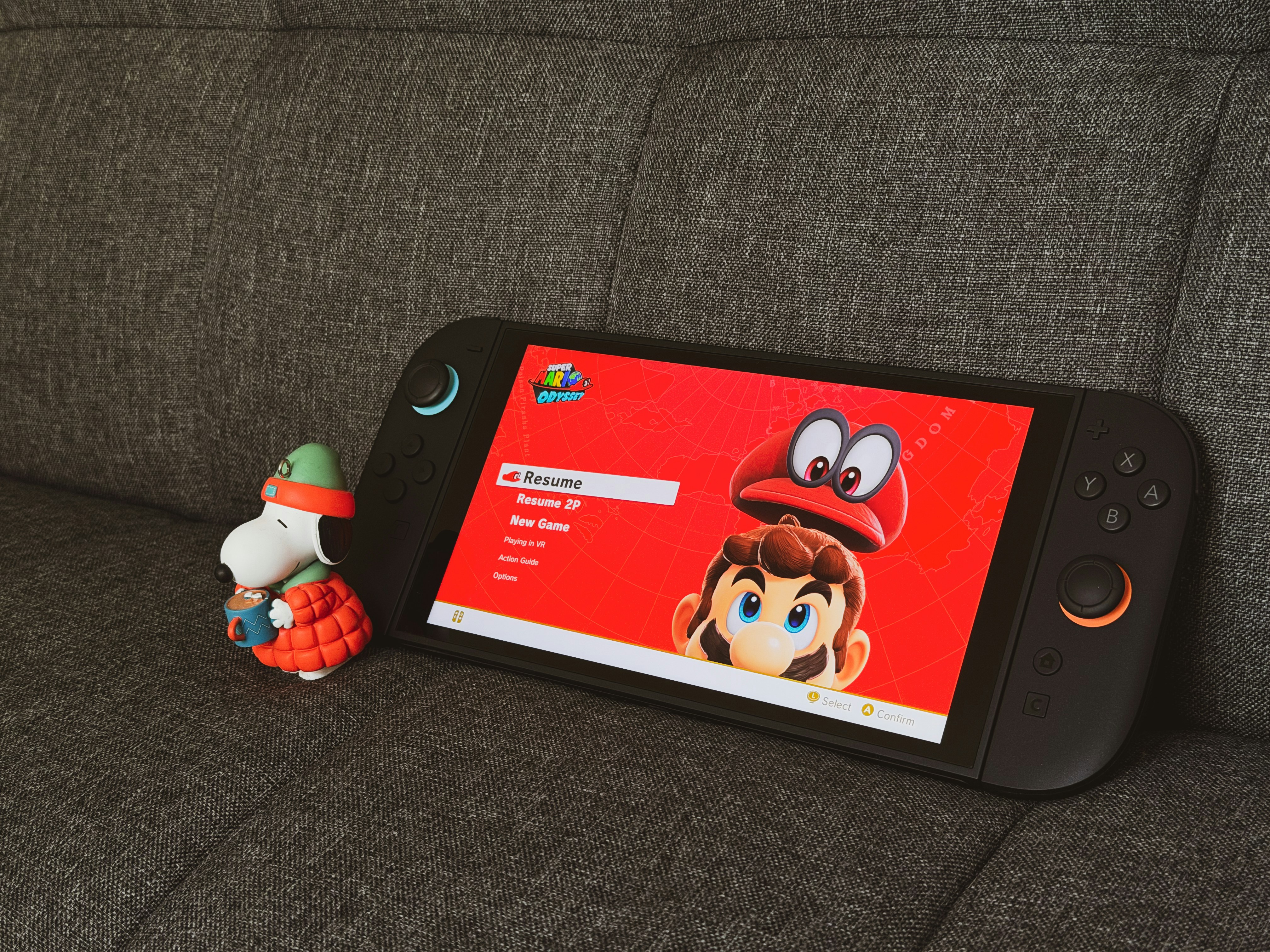 A Nintendo Switch in handheld mode rests on a couch with a small Mario figurine beside it.