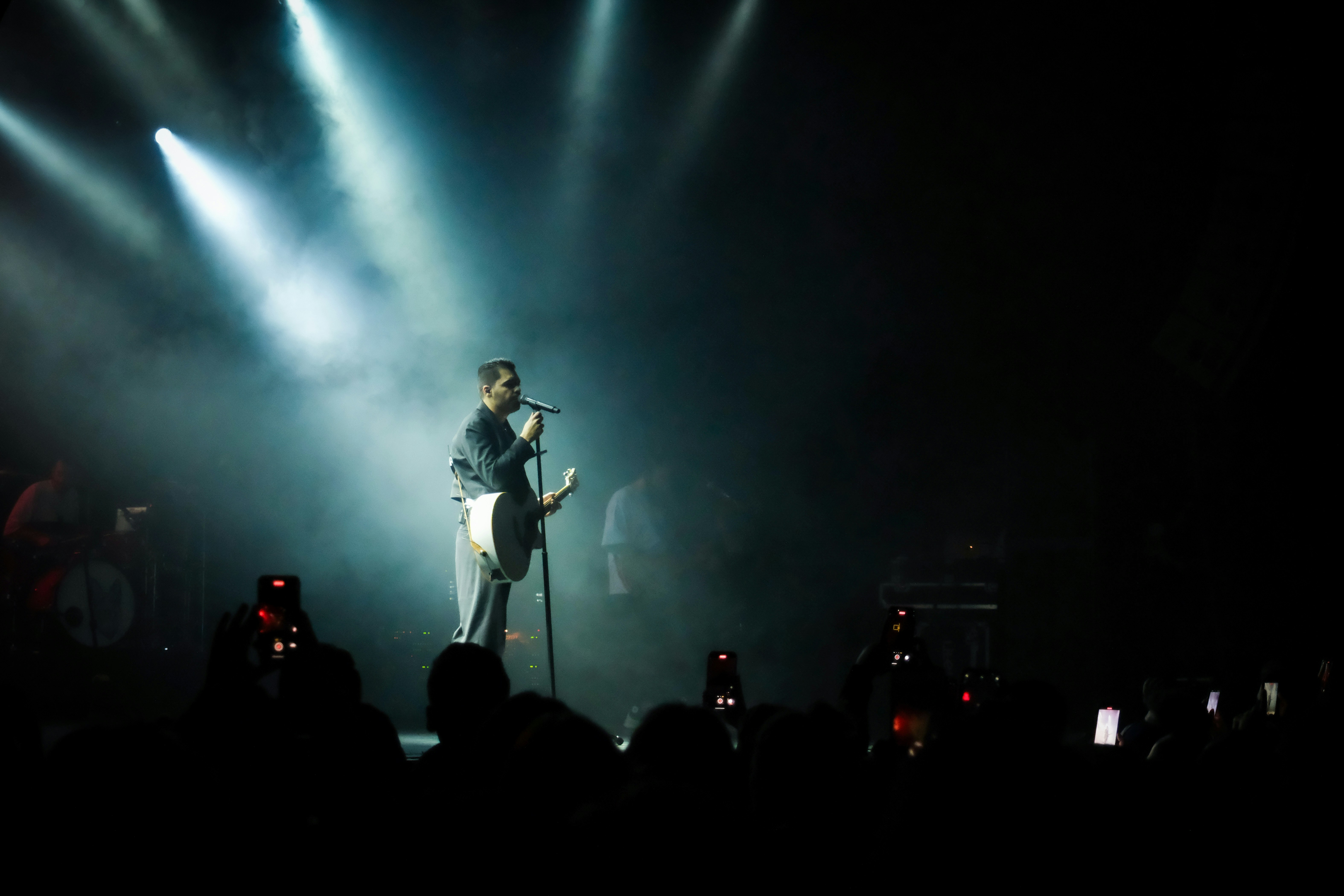 Singer on stage with guitar under dramatic lighting, silhouettes of the audience, and phones flickering in the foreground.