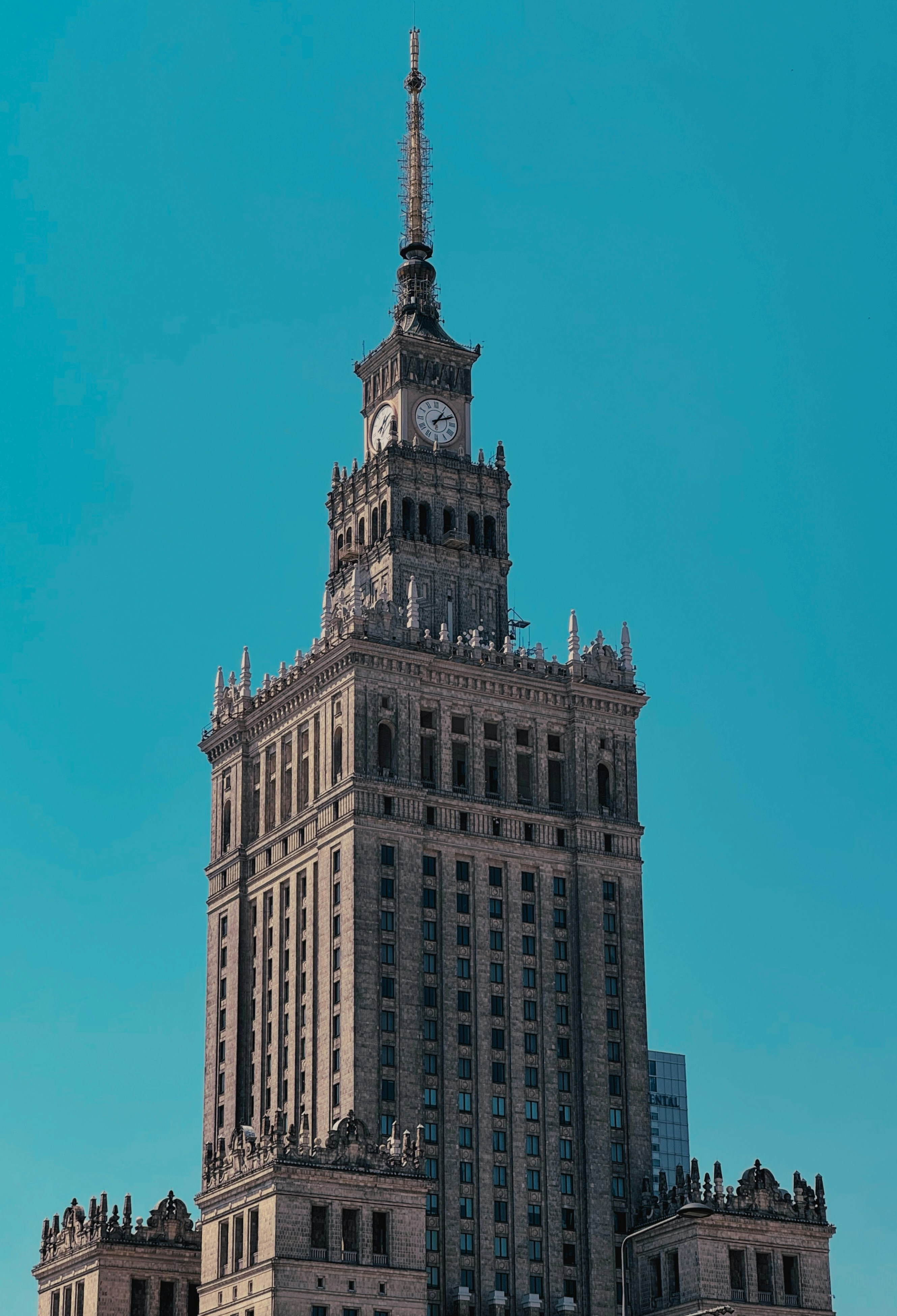 Clock-taced tower rises above a blue sky, detailing ornate architecture and spire. Prominent clock face sits near the top, emphasizing vertical grandeur of the historic structure.