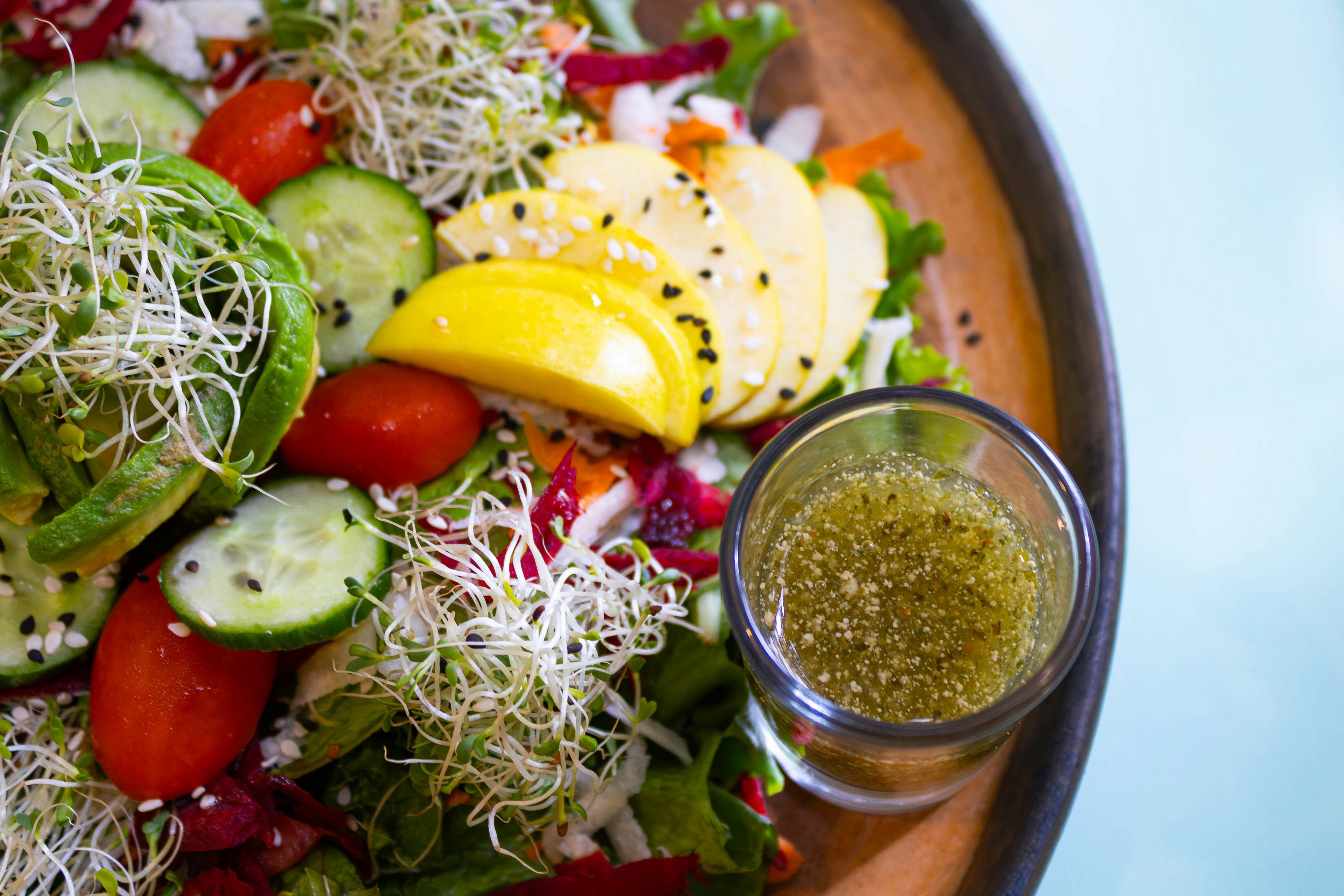 Colorful salad with cucumber, tomato, avocado/greens and scattered sprouts, accented by a sliced lemon and a small glass of dressing on a wooden plate.