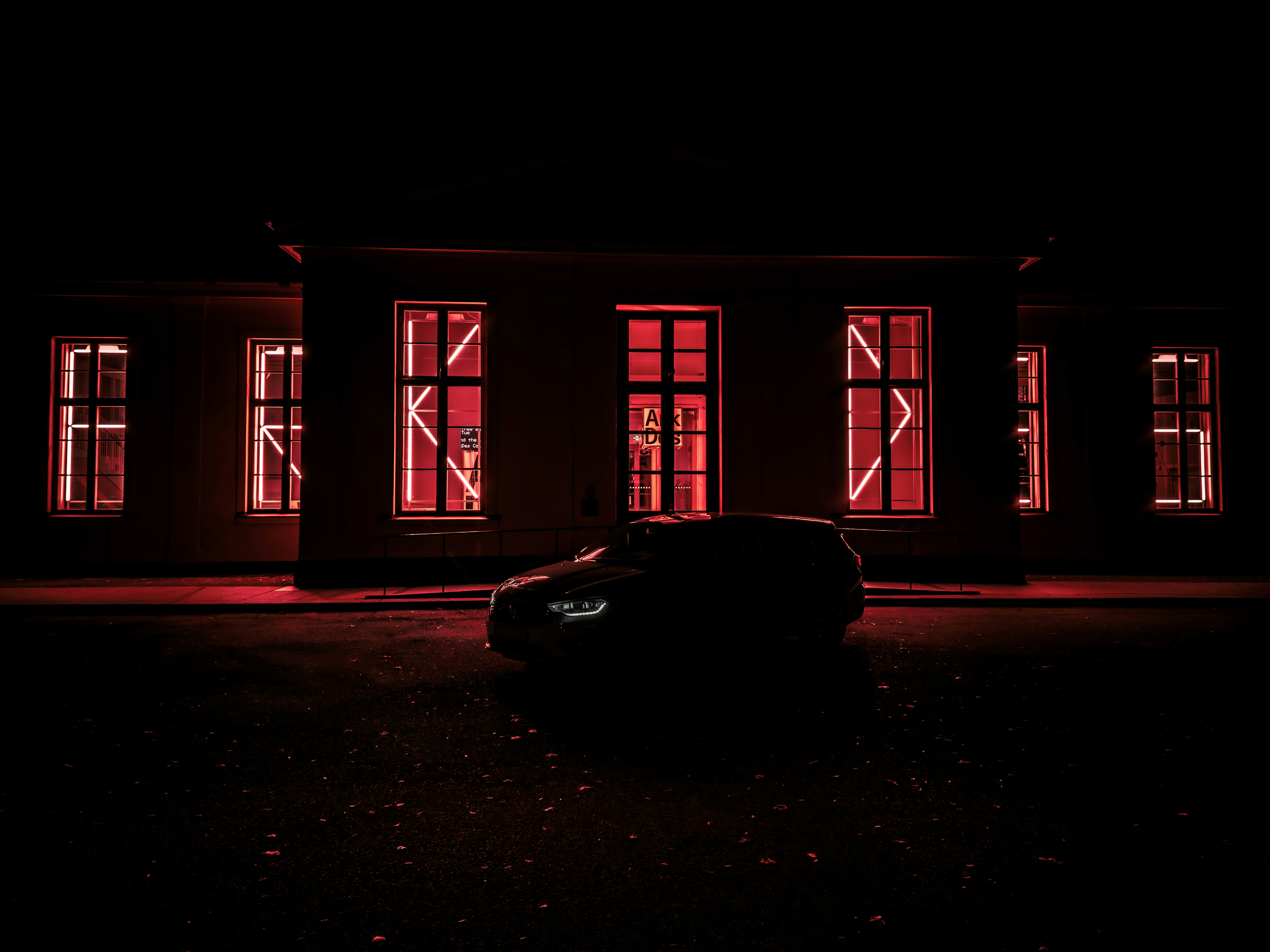 A dark urban scene features a car parked in front of a building glowing with red neon window outlines, creating a moody, cinematic silhouette.