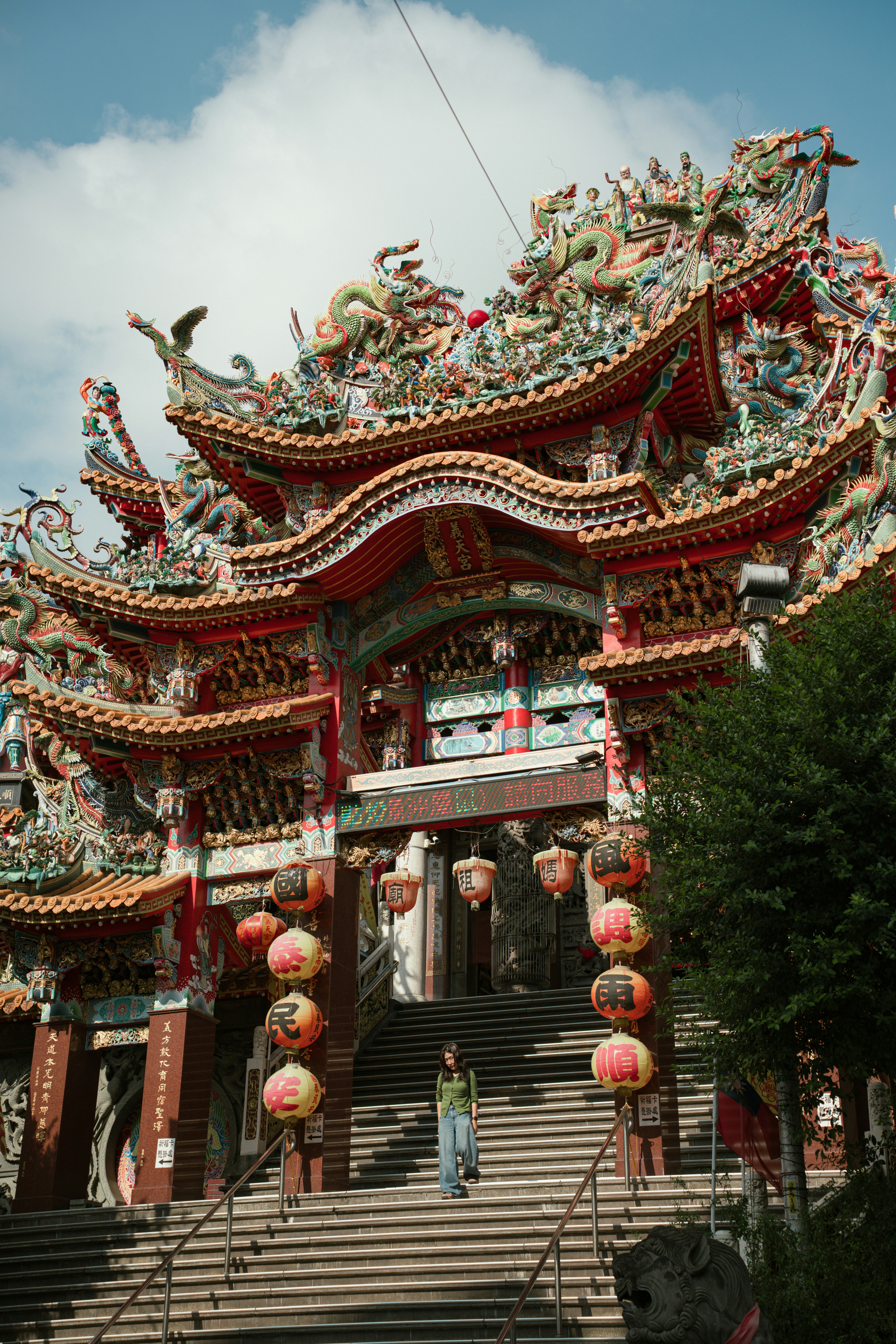 Colorful ornate temple façade with dragon motifs and red lanterns guarding stone steps; a lone visitor stands at the entrance, framed by the temple's intricate carvings.