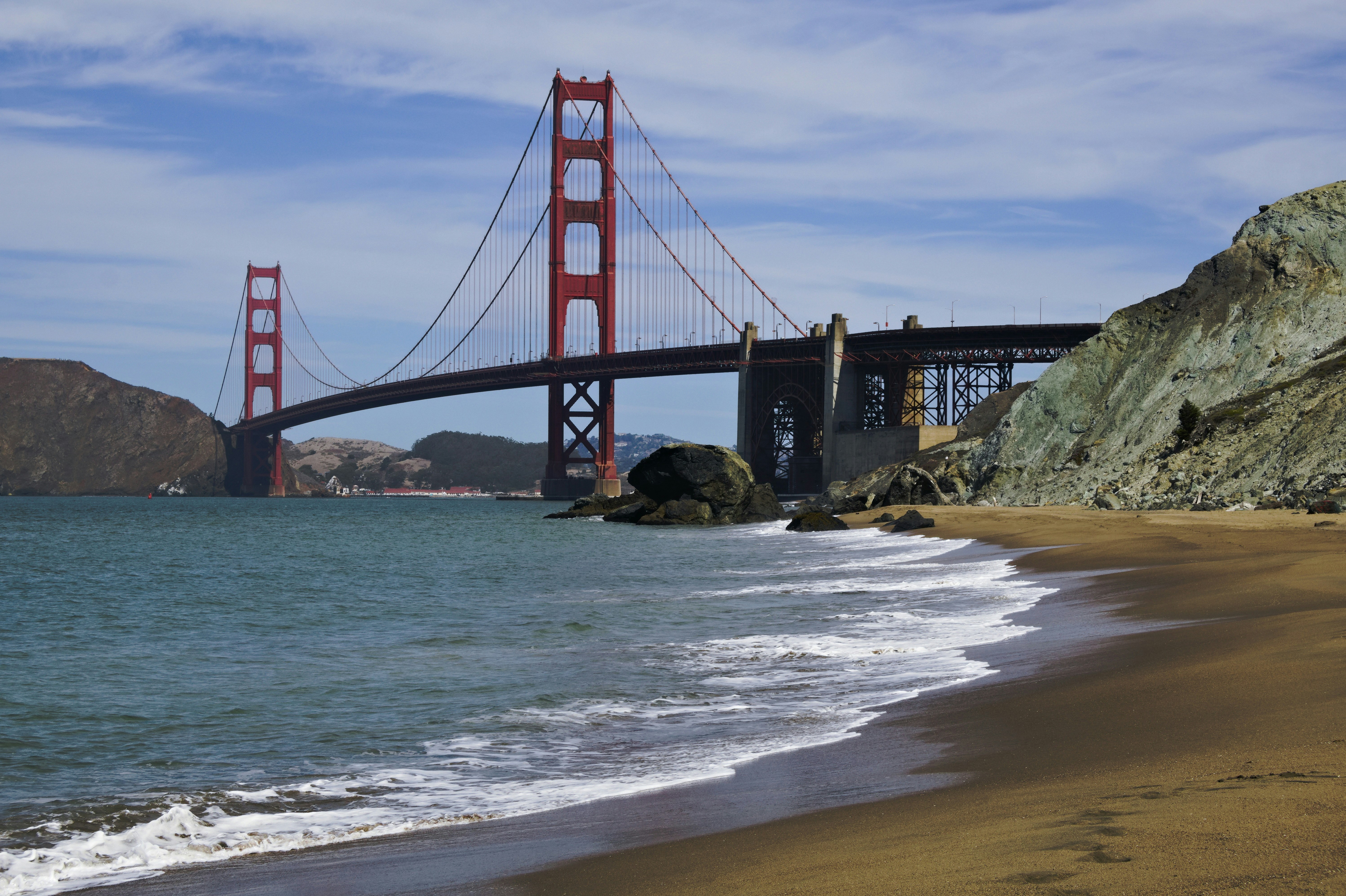 Golden Gate Bridge visible across sandy shoreline with waves in foreground and rugged rocks to the right. A coastal scene emphasizing coastal landscape and iconic bridge silhouette.