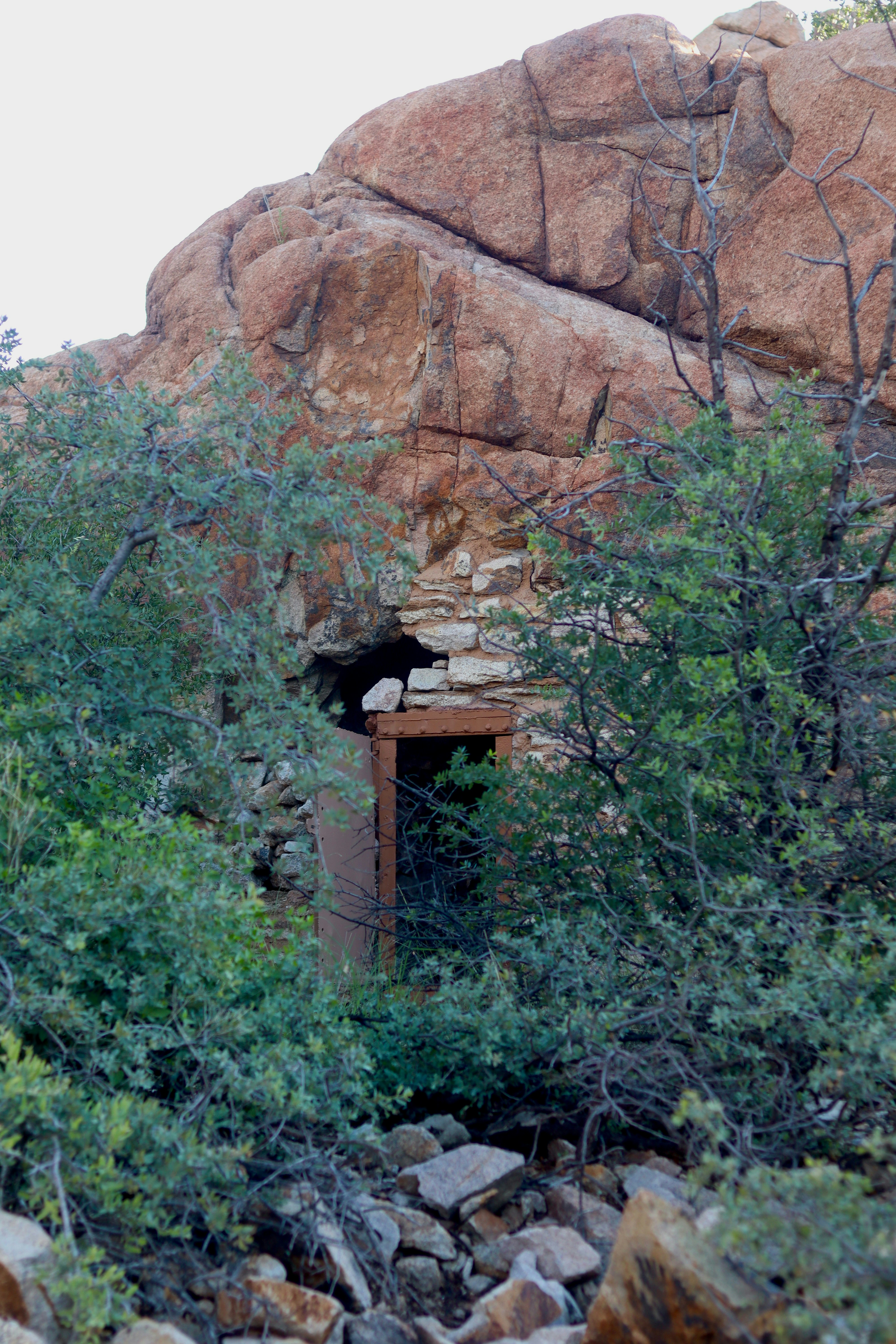 Overgrown rock facade reveals a weathered doorway framed by rugged boulders and scrubby vegetation.