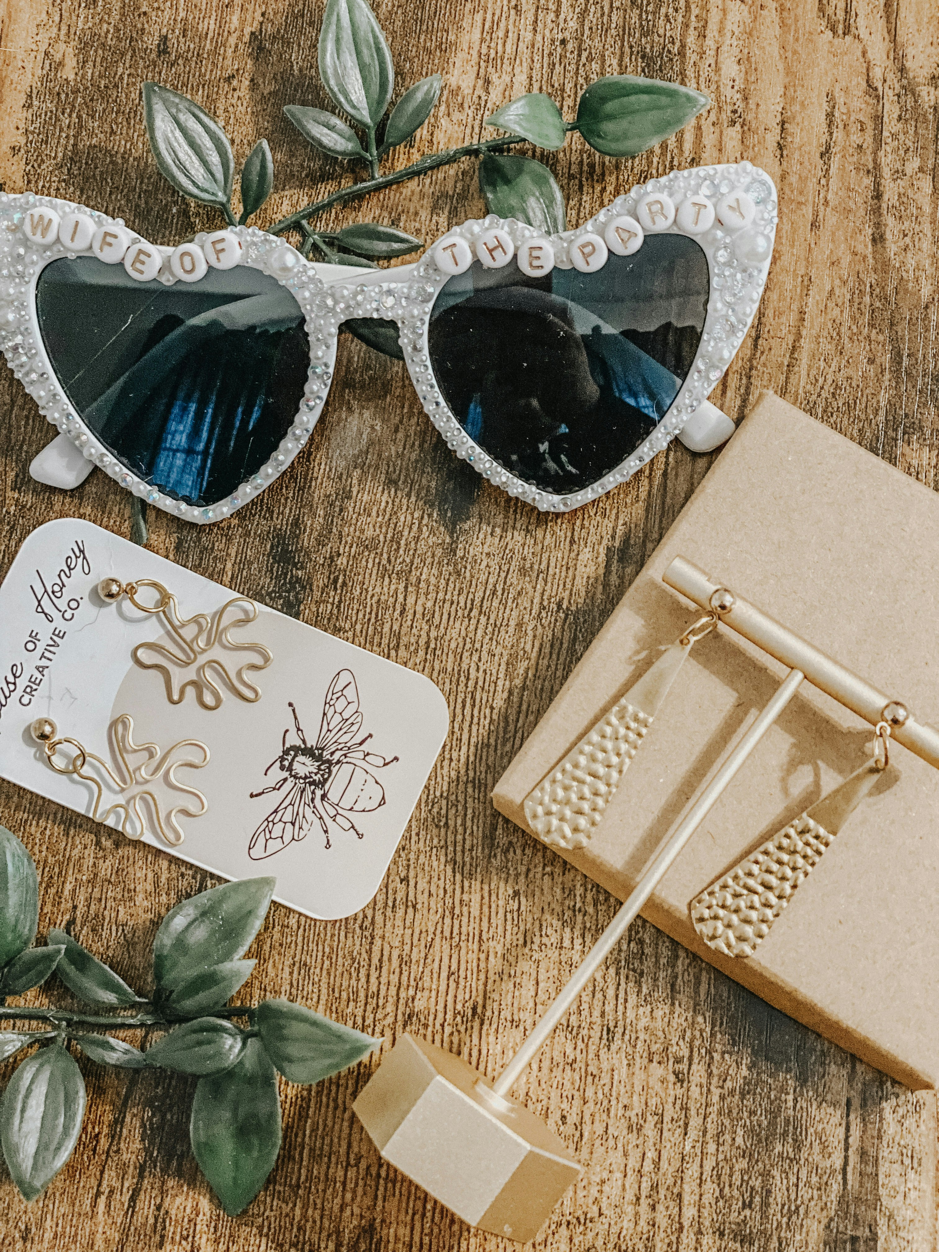 Pearl-trimmed heart sunglasses and gold jewelry lay on a warm wooden surface, with a small wrapped gift and plant accents adding to a cozy, craft-chic display.