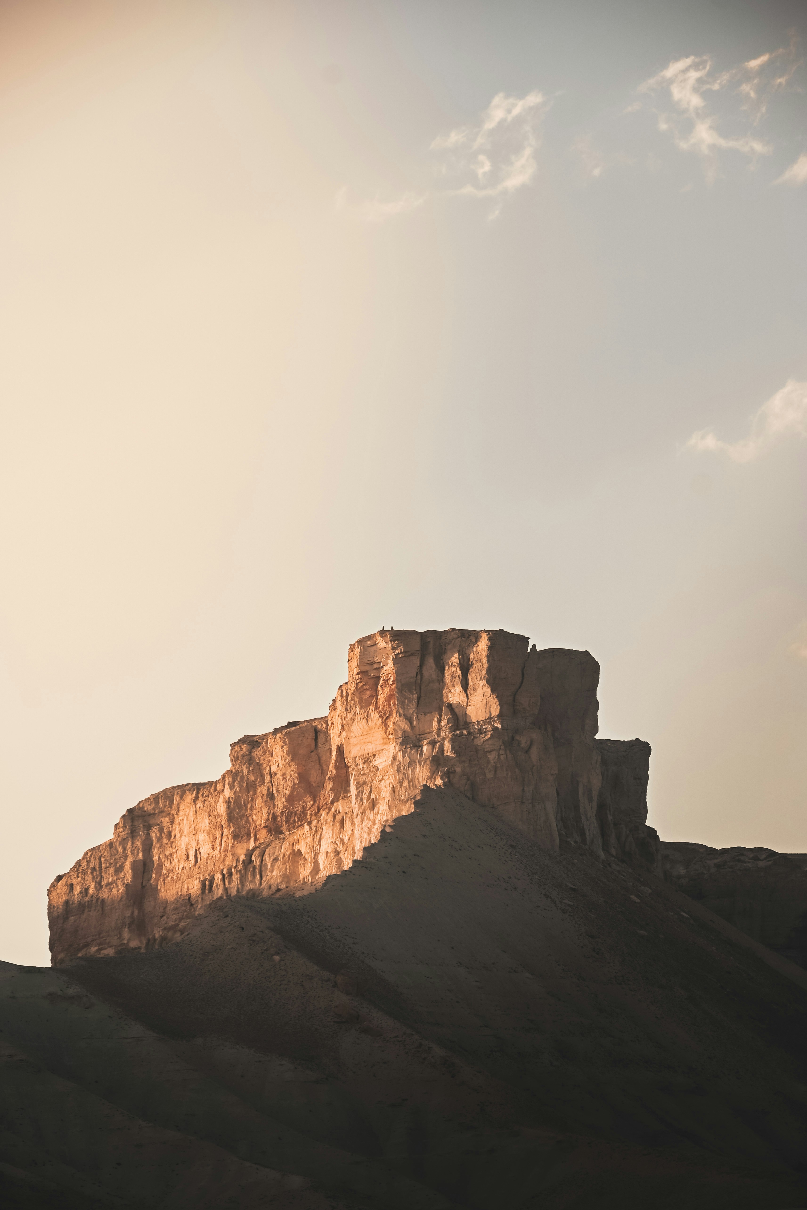 Warmly lit rock formation rises from shadowed slopes against a pale sky, emphasizing its rugged silhouette and soft evening light.