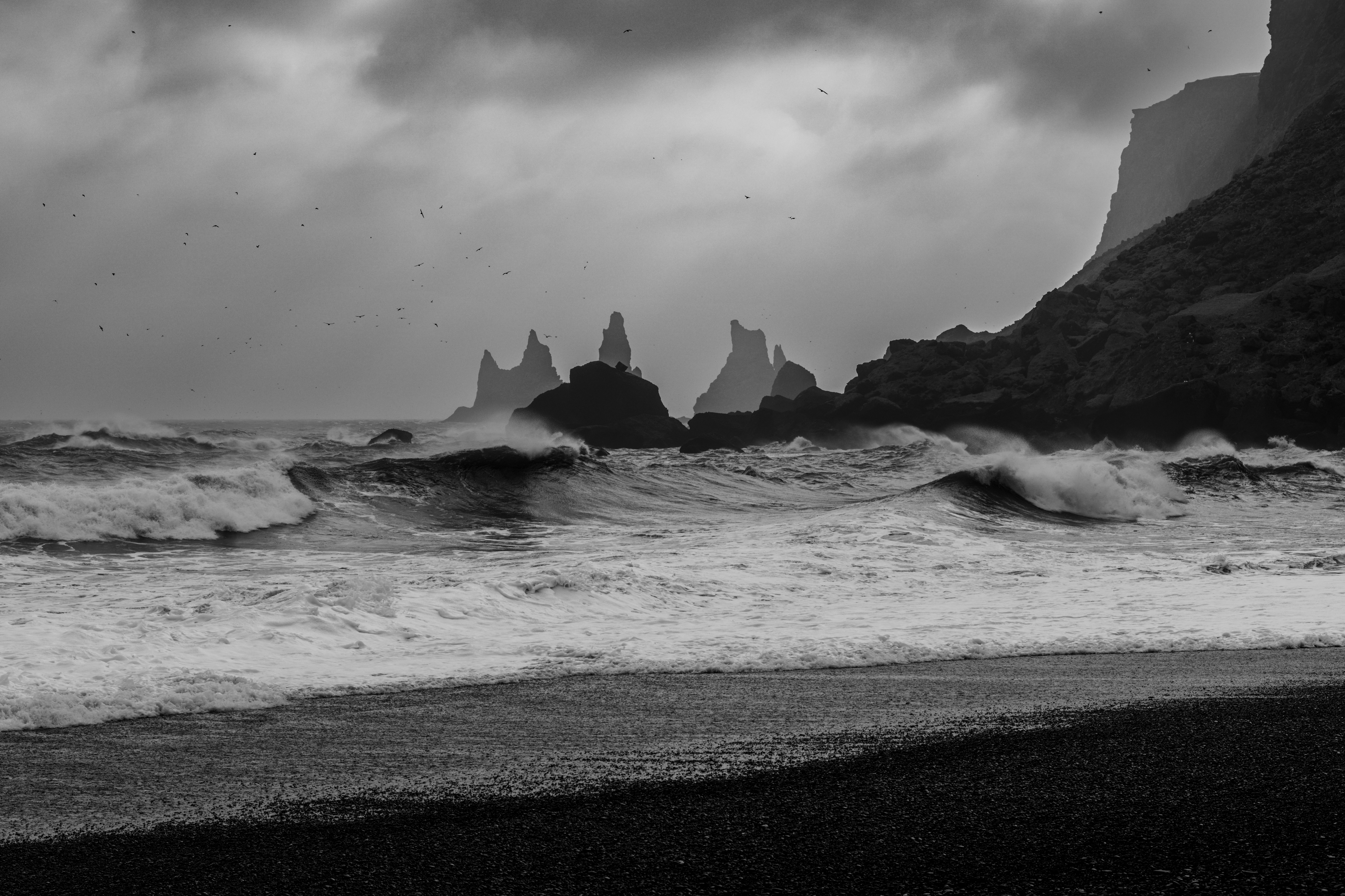 Monochrome coastal scene with jagged rock formations rising from the sea and waves breaking on a pebbled shore; a moody, atmospheric seascape emphasizing texture and contrast.