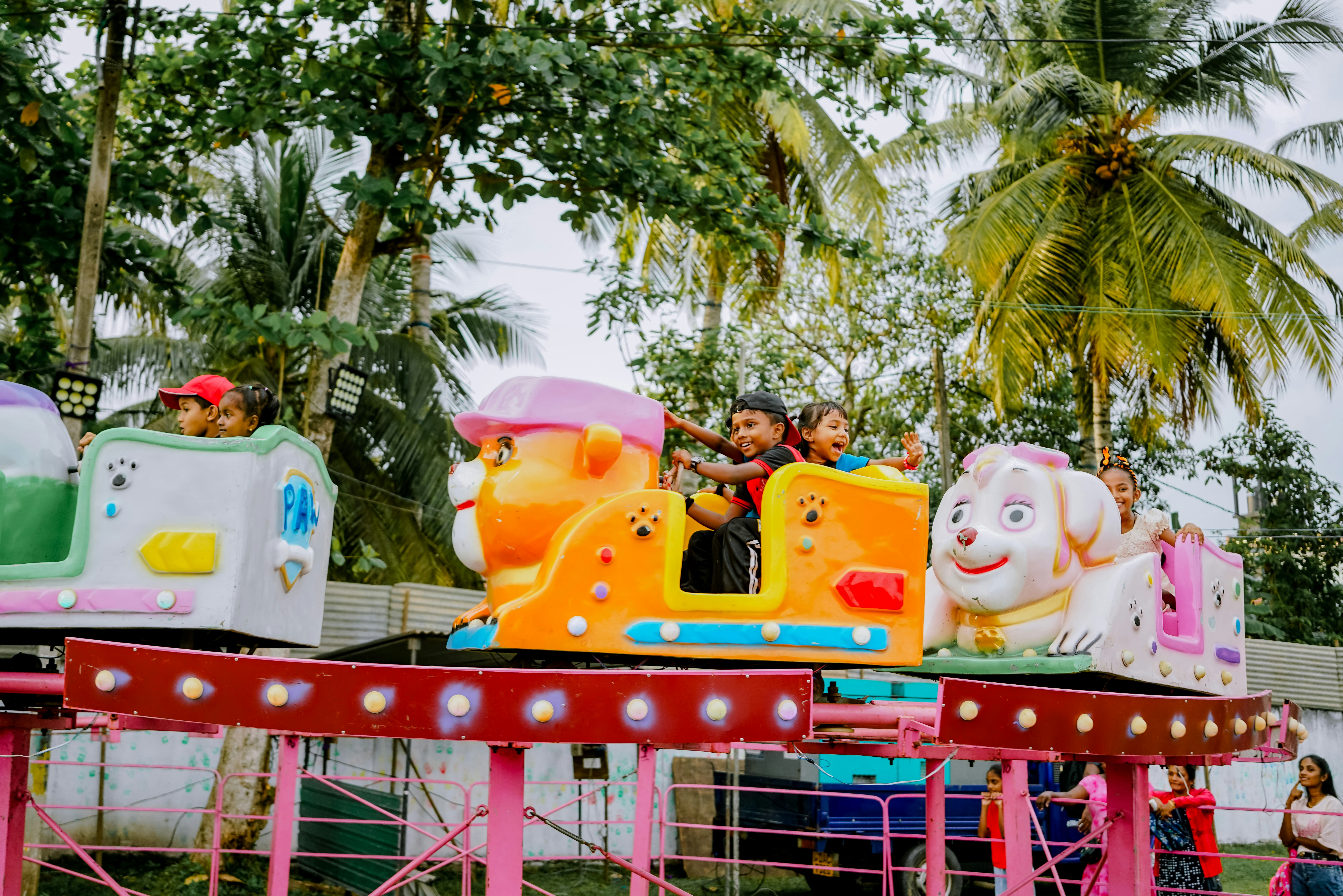 A vibrant amusement ride with cartoon animal carriages carries children against a backdrop of palm trees and a bright sky.