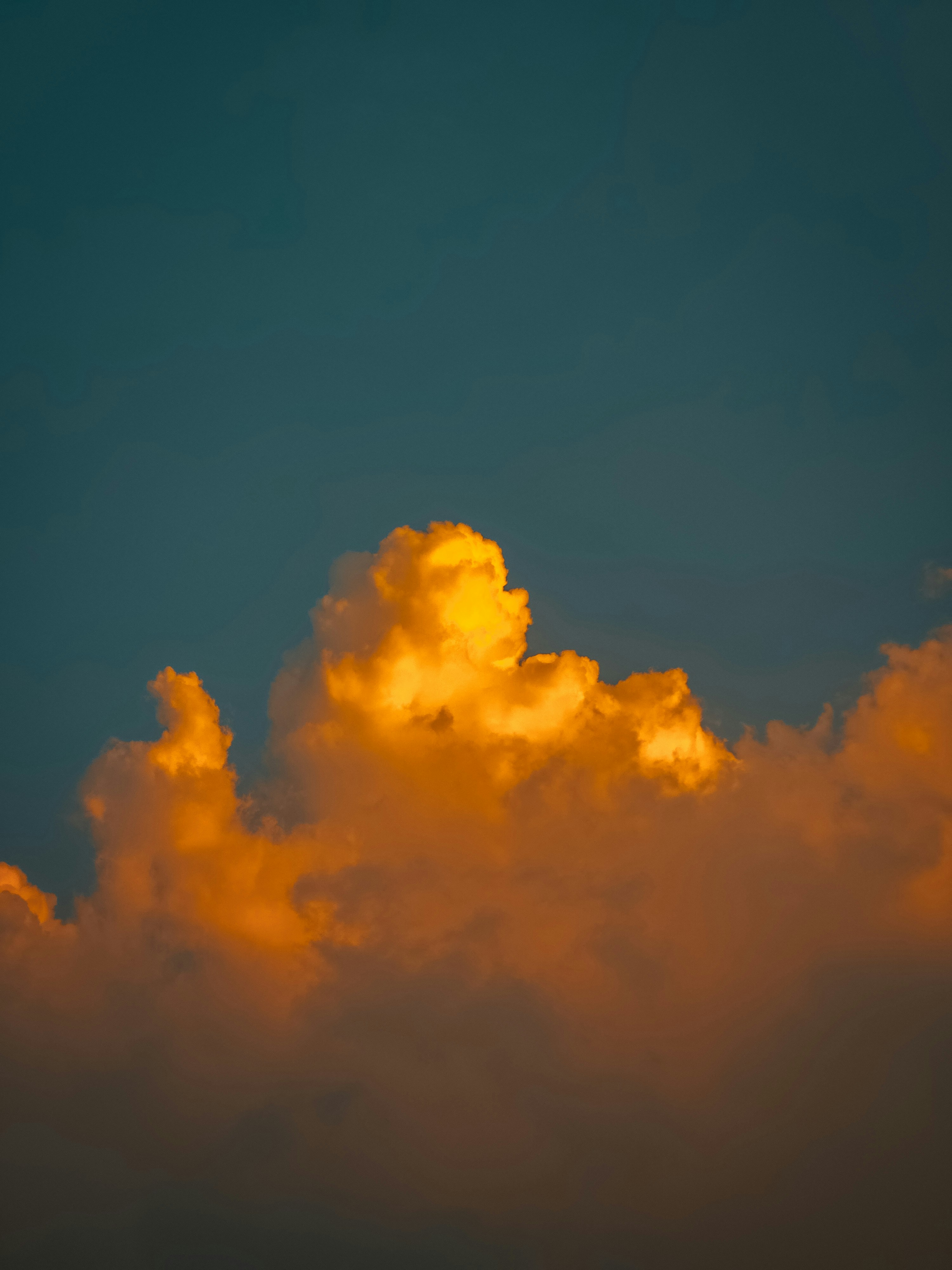 Vibrant orange cloud formation rises against a deep teal sky, emphasizing a dramatic sunset glow and atmospheric depth. Aerial cloudscape with warm highlights dominates the frame.