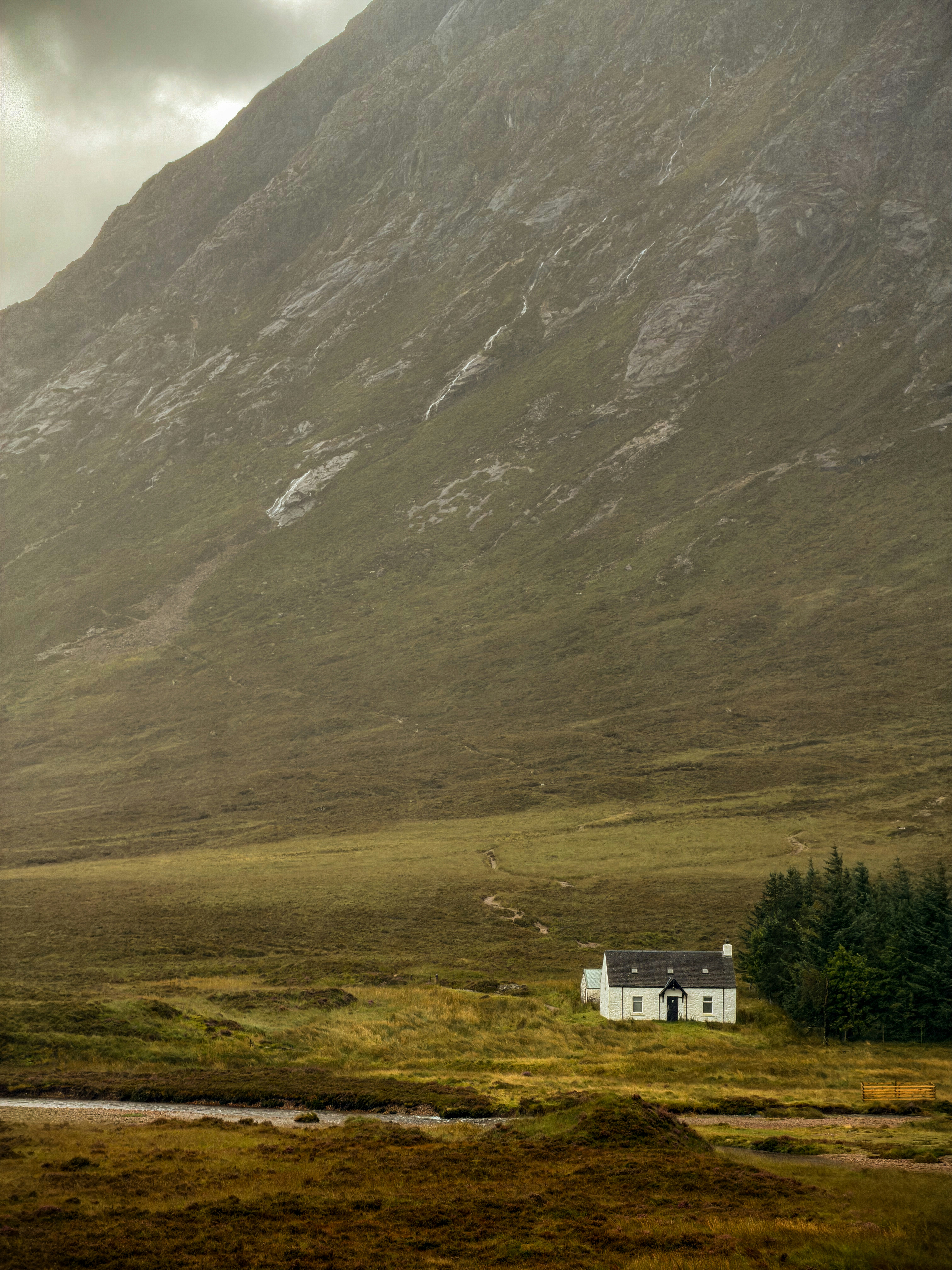 A solitary white cottage rests at the foot of a towering, rugged hillside, with a narrow path winding toward it across a moorland landscape.