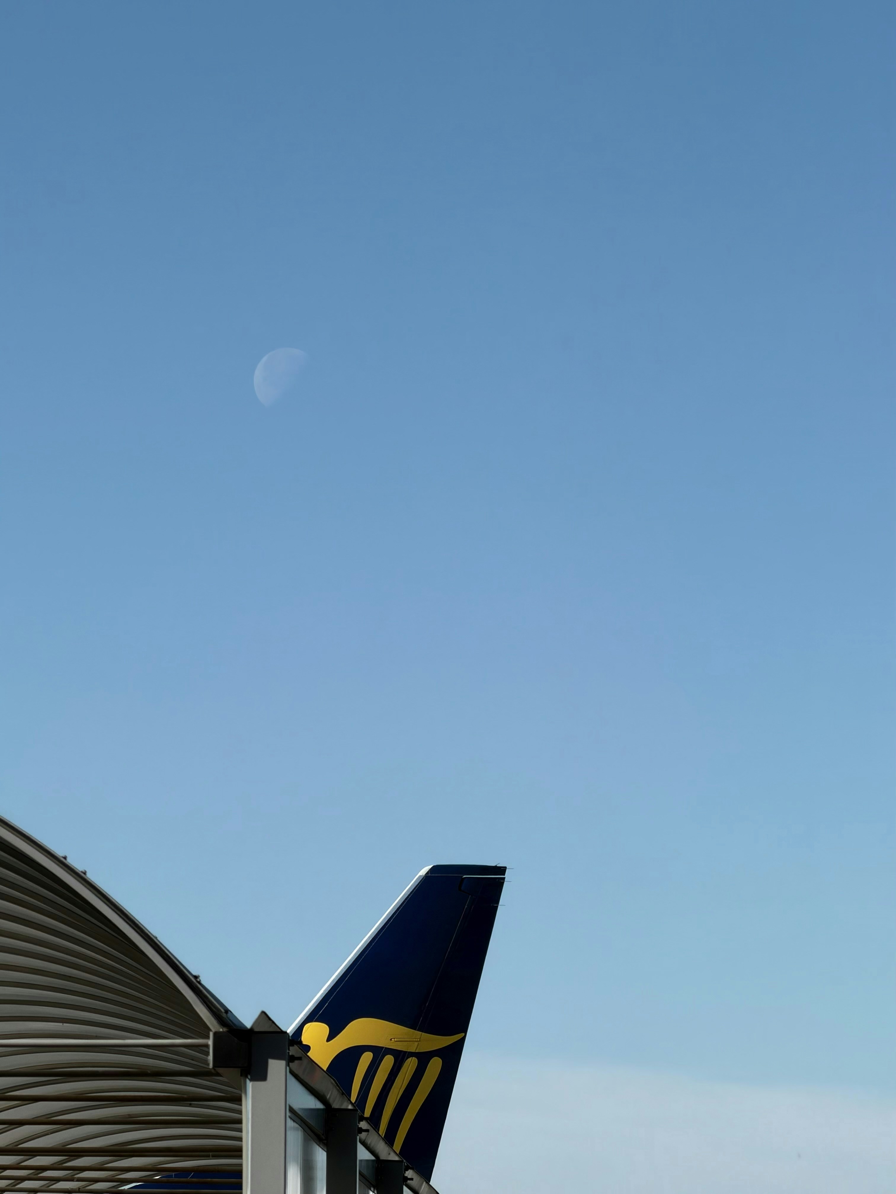 The tail fin of a Ryanair airplane against a clear blue sky with a visible moon