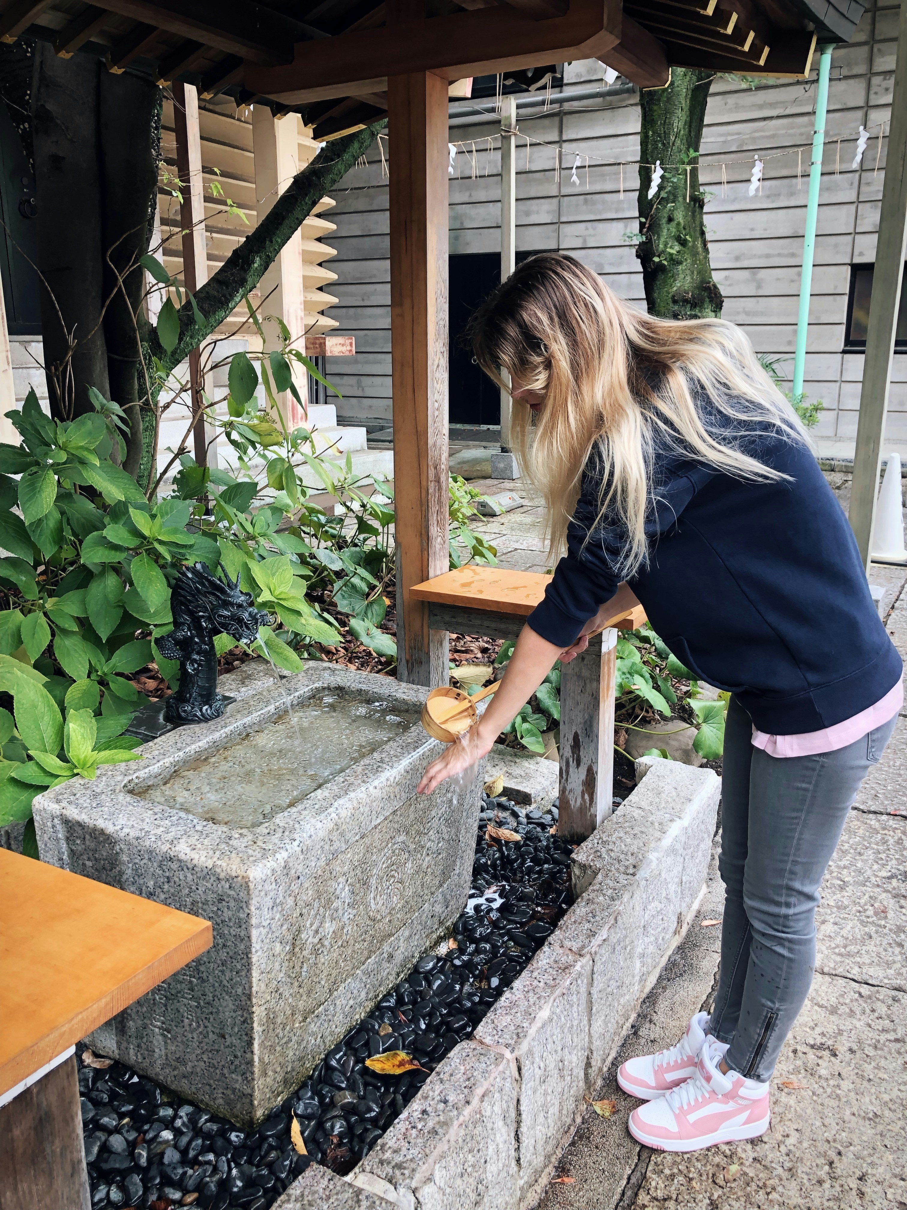 A woman using a wooden ladle to wash her hands at a stone fountain