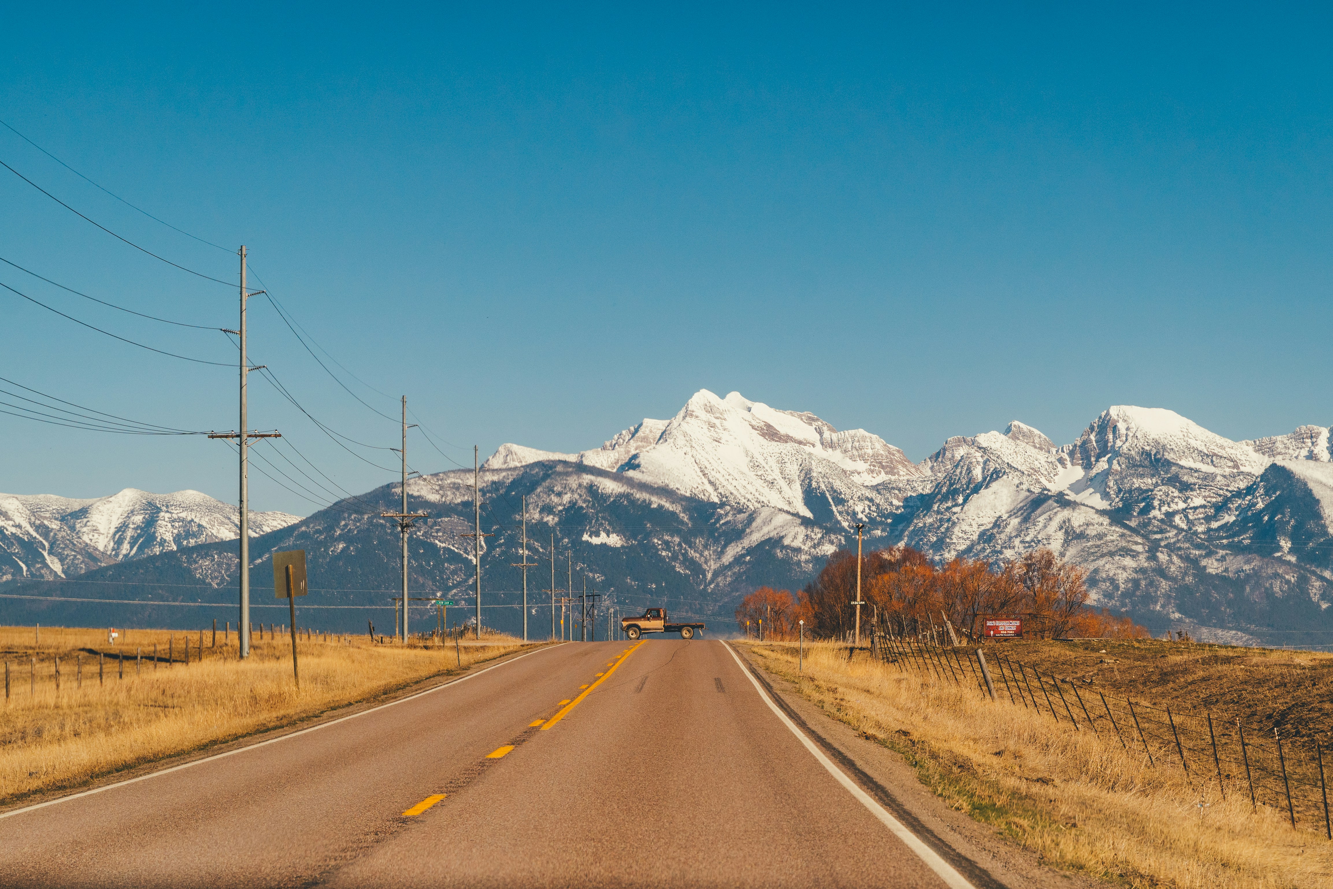 A straight rural highway stretches toward a snow-capped mountain range under a clear blue sky, with fields flanking the road and utility poles receding into the distance. The scene captures open landscape and distant alpine scenery.