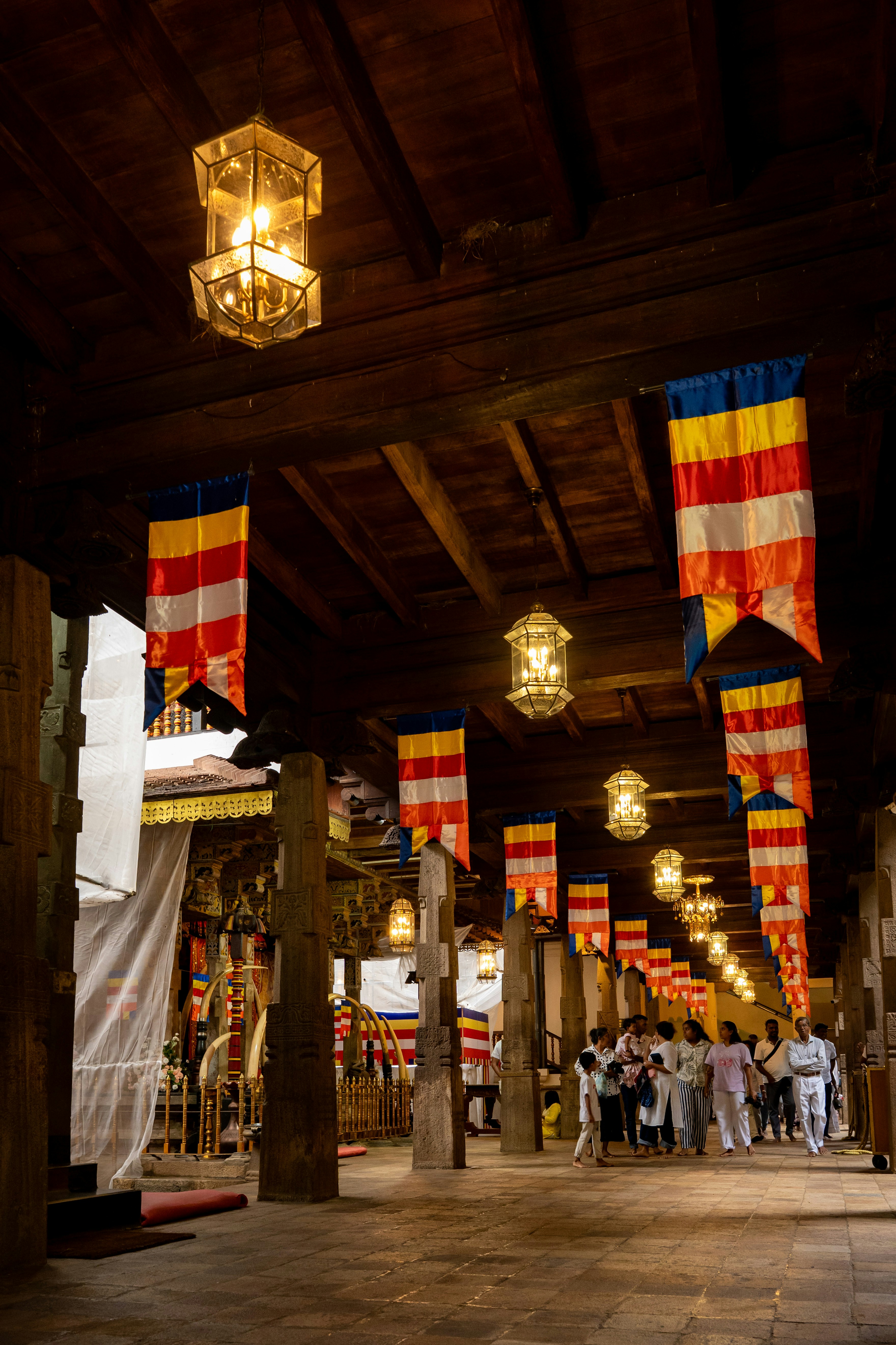 Warm-lit temple corridor with hanging lanterns and vertical color-striped banners; a group of people gathered toward the far end, suggesting a cultural or ceremonial gathering.