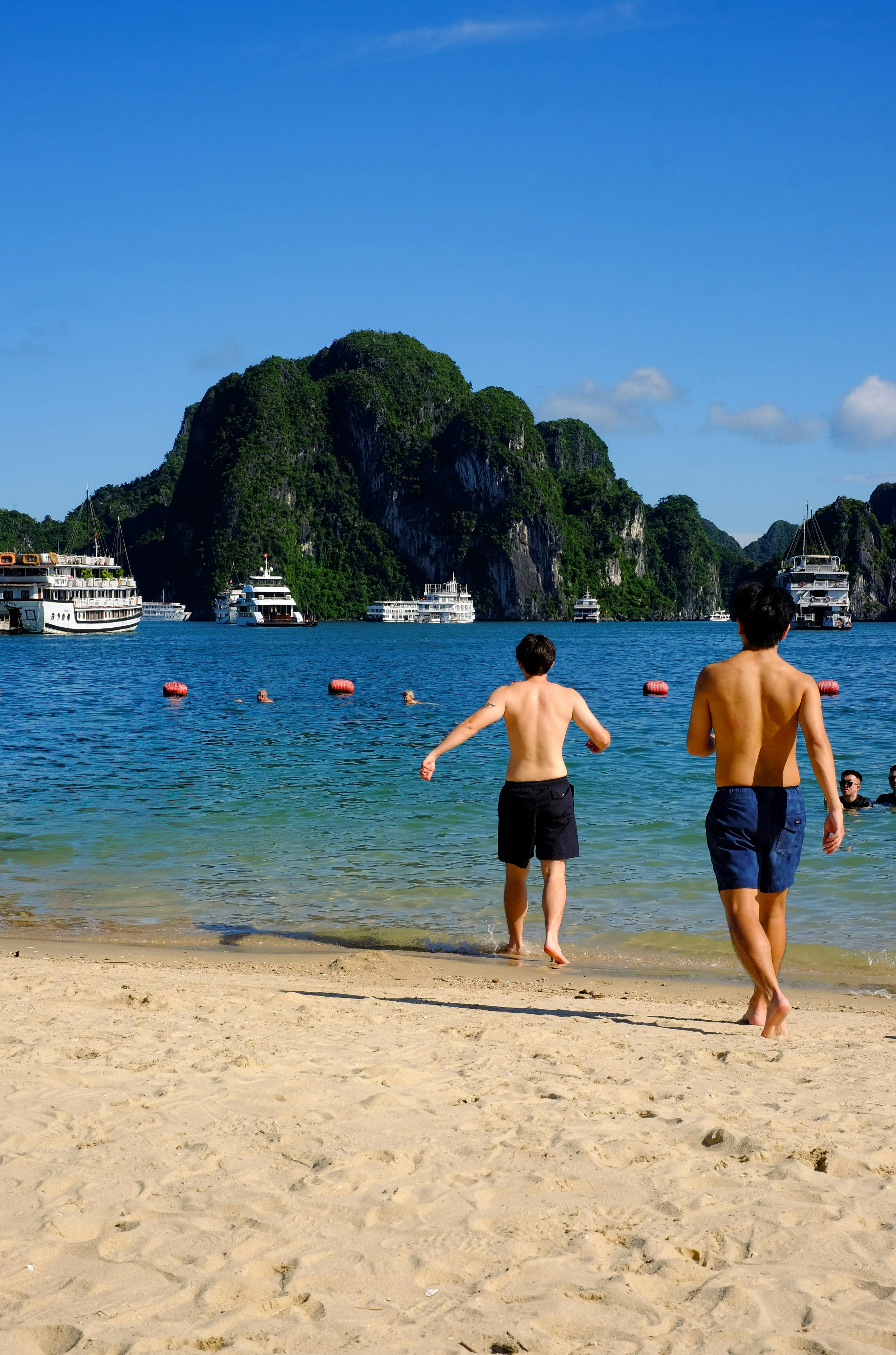 Two shirtless friends sprint toward the turquoise surf as limestone islets rise beyond a busy bay