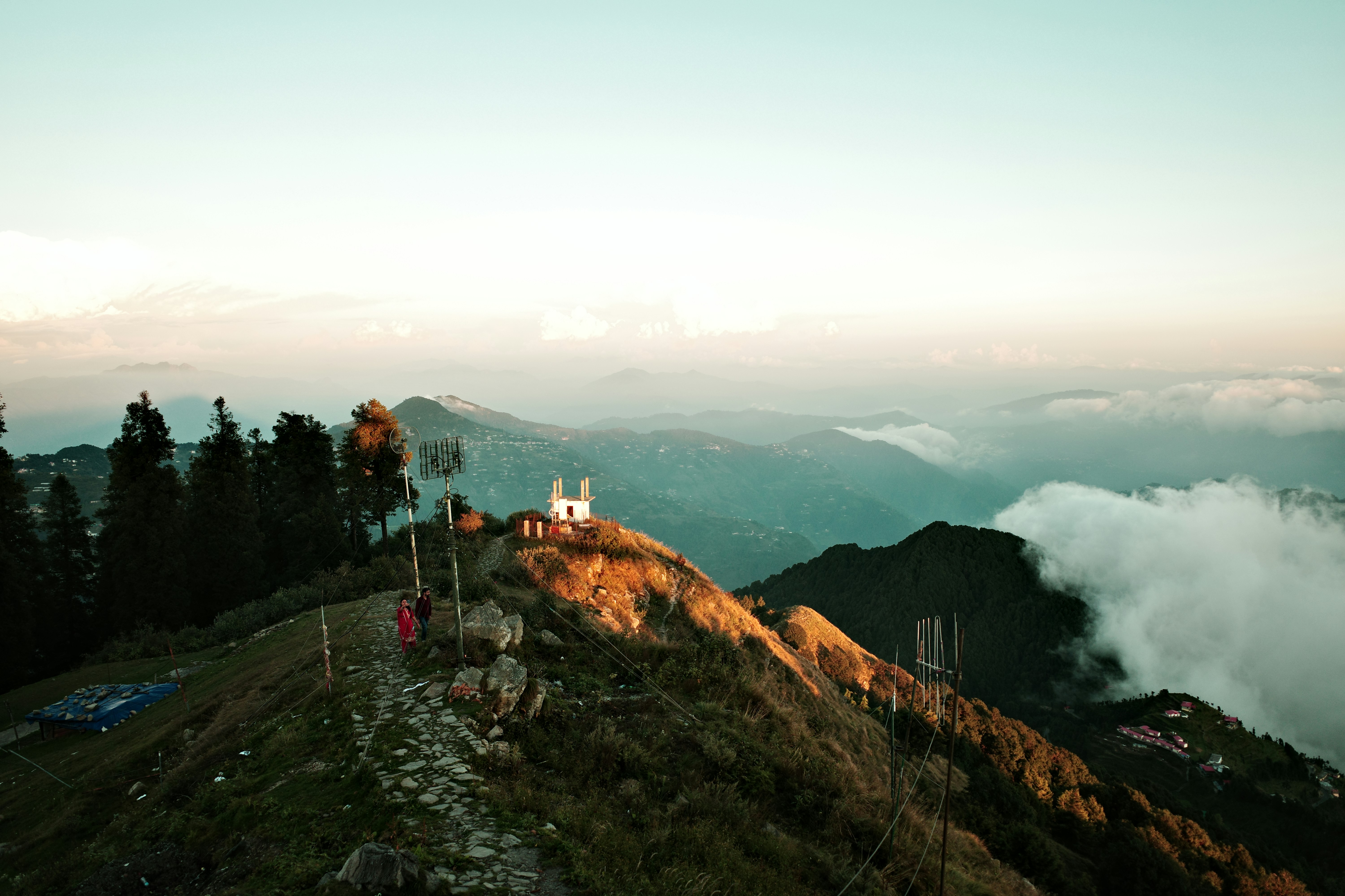 Ridge path leads to a small structure atop a hill with a distant chapel, against rolling mountains and a sea of clouds; a tranquil landscape scene.