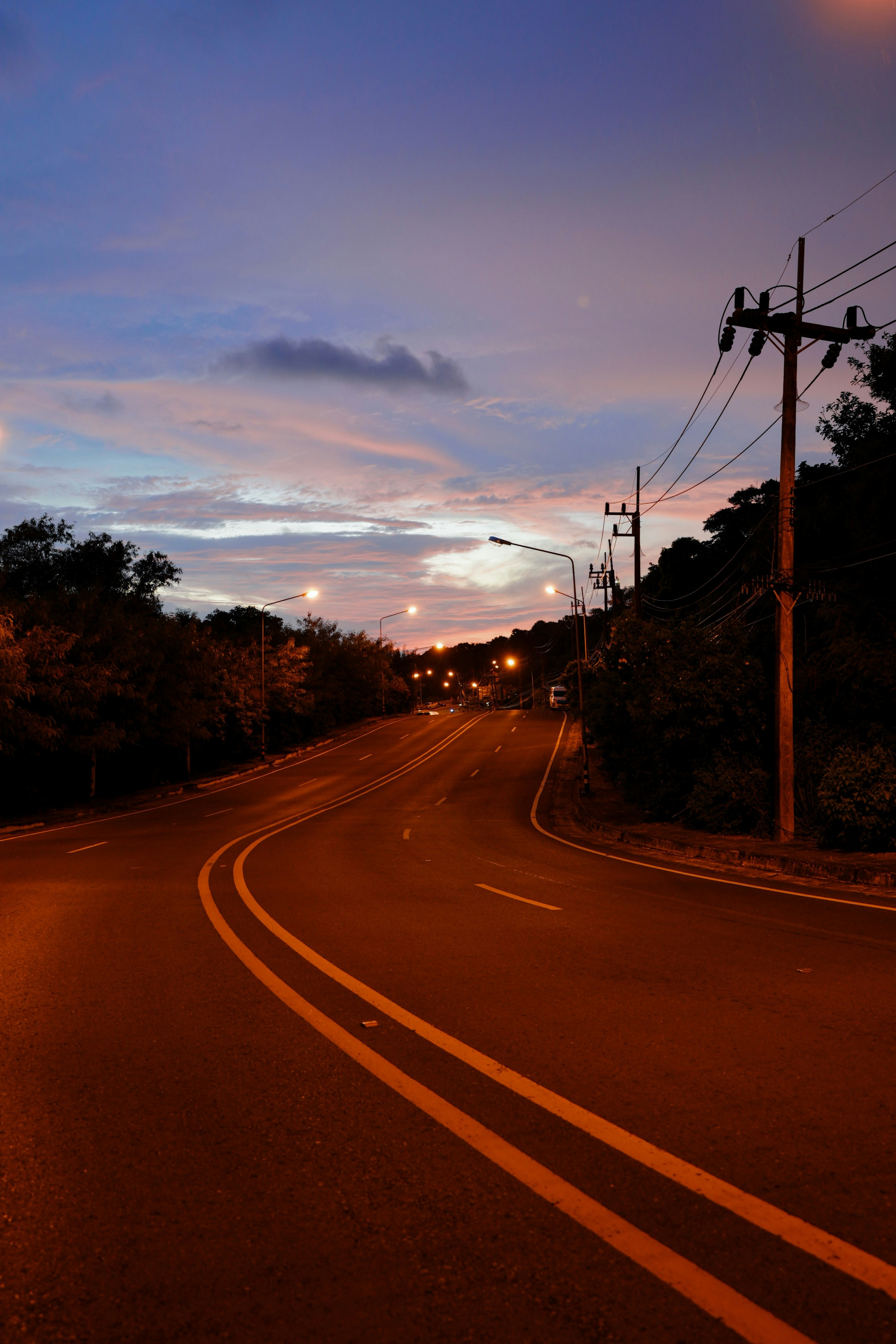 A winding empty asphalt highway illuminated by streetlights during a beautiful twilight sunset.