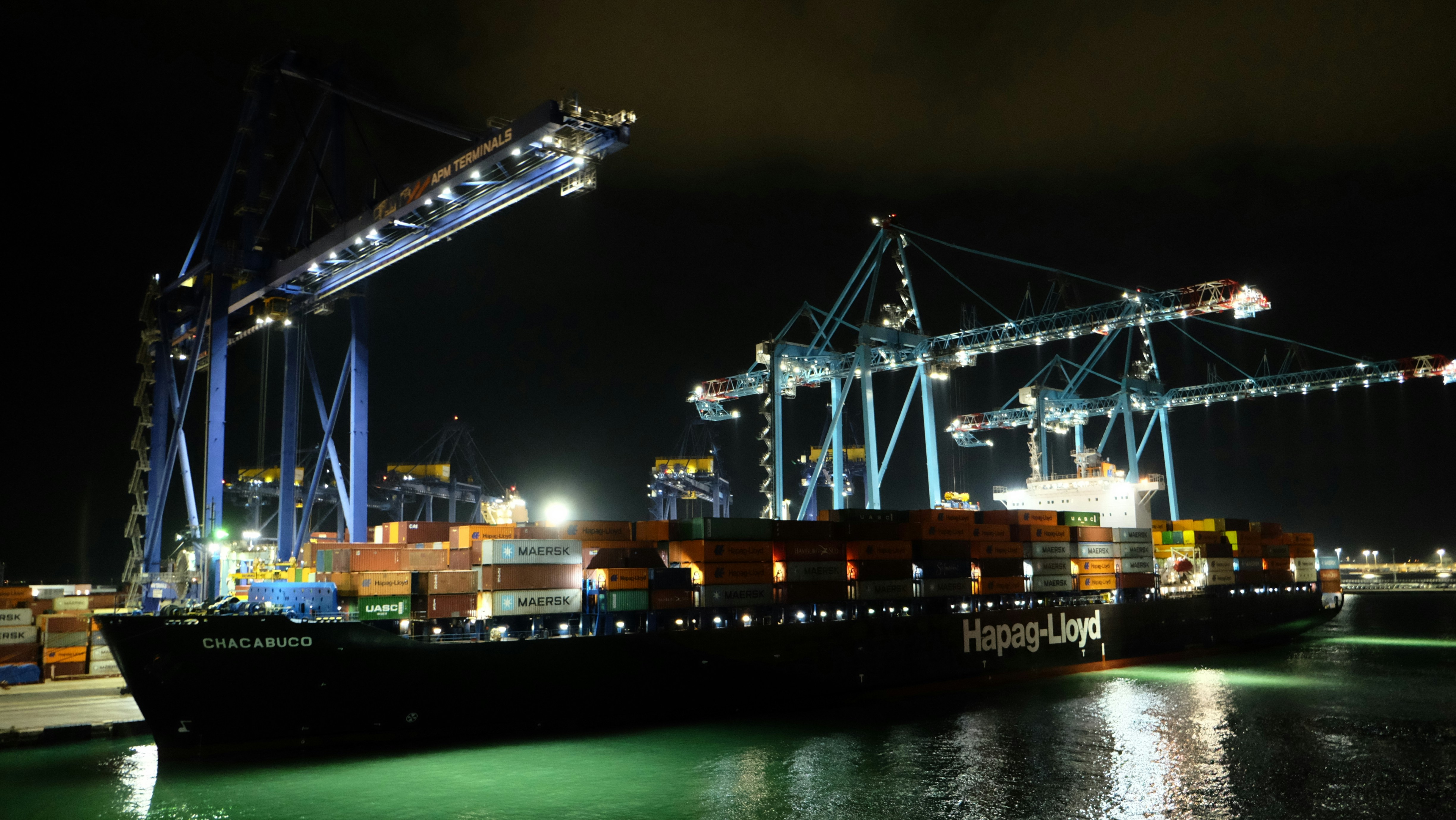 Container ship docked at night under tall gantry cranes, illuminated by port lights with greenish water reflections.