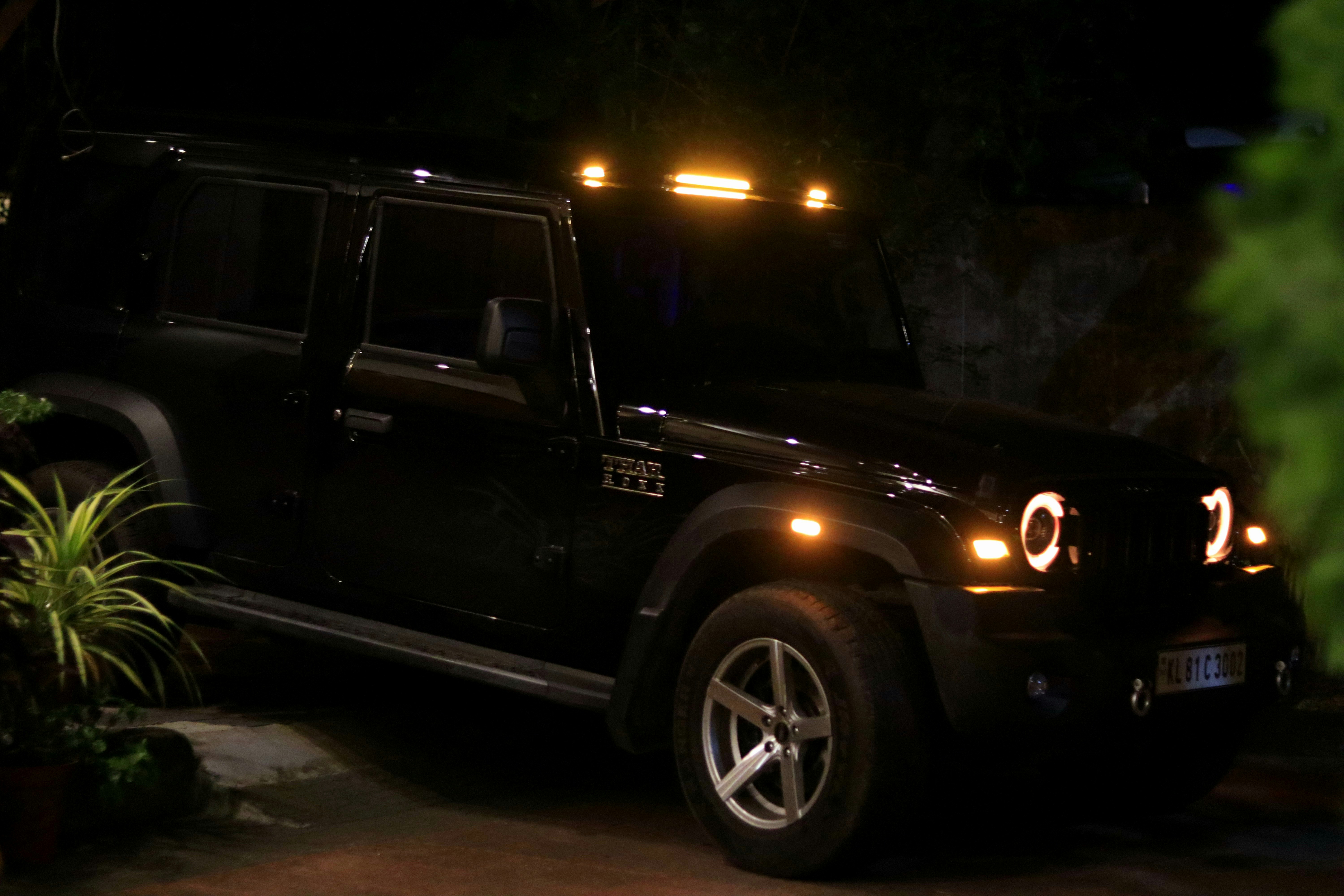 Dark-toned SUV parked at night with illuminated roof lights and front headlights dimly glowing, hinting at a nocturnal driveway scene. The vehicle is the focal point, partially framed by foliage on the left.