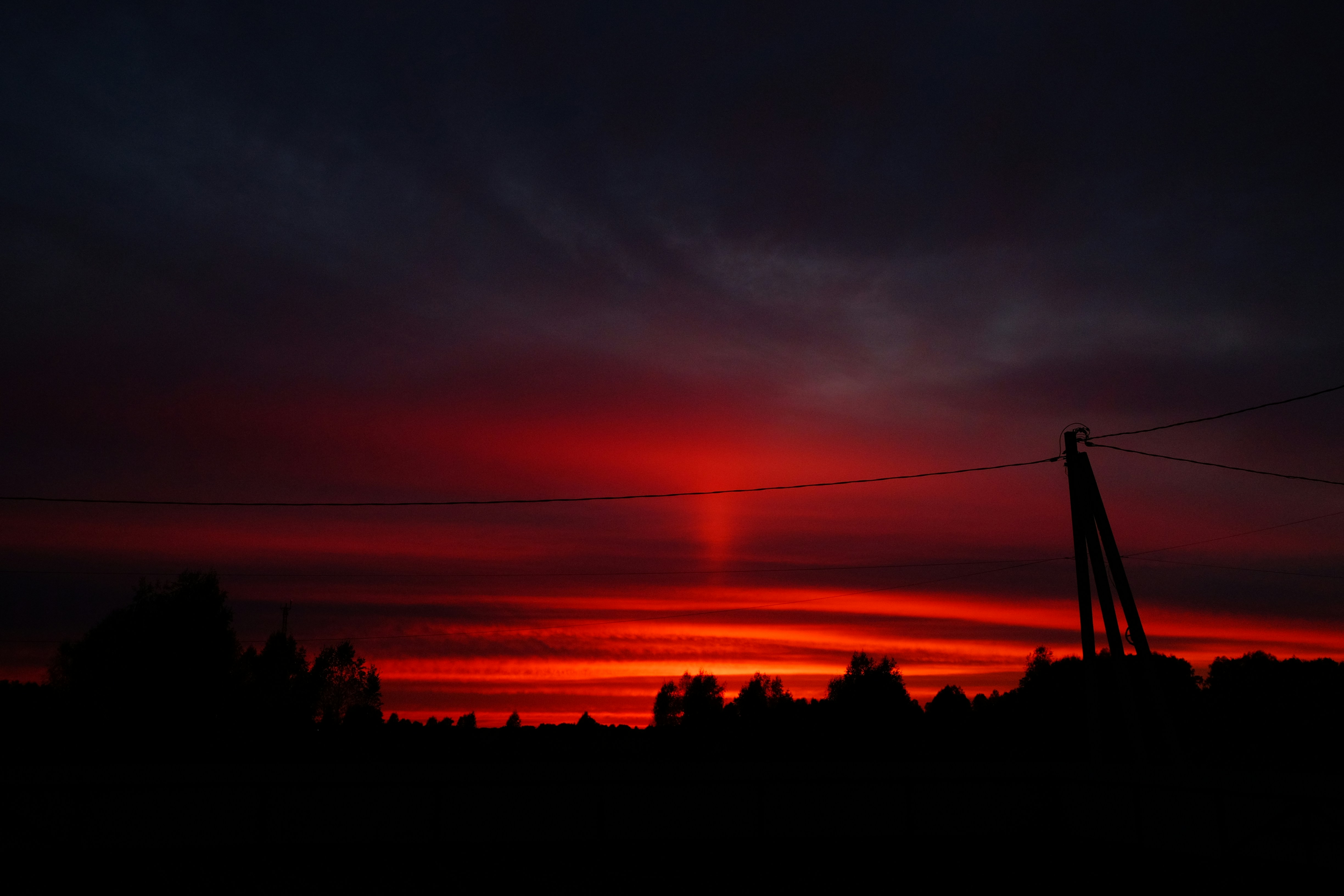 Silhouetted landscape under a deep red sunset with a lone utility structure crossing the frame, foreground dark and unlit.