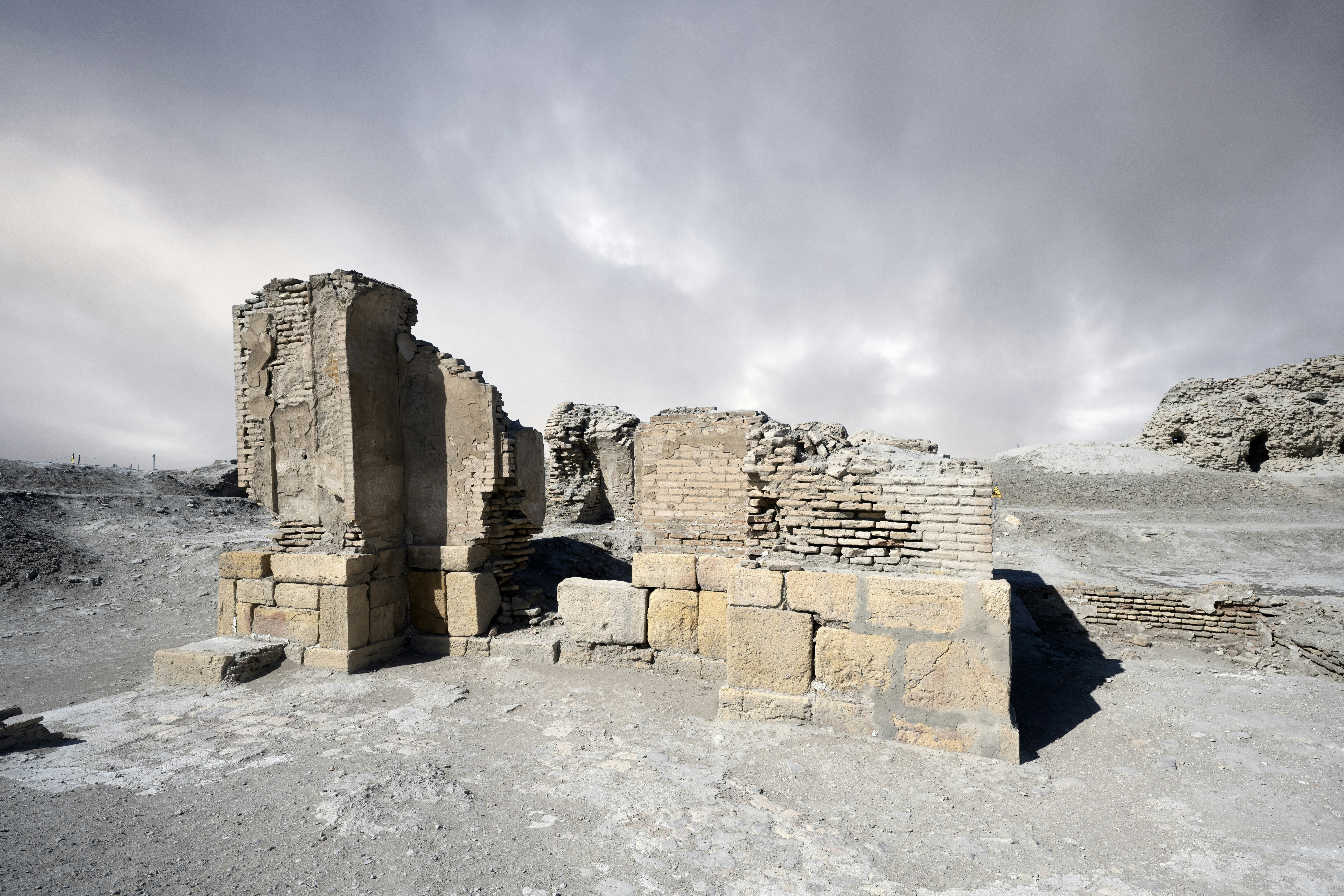 Ancient stone structures rise from a dusty, sunbaked landscape under a dramatic, cloud-filled sky.