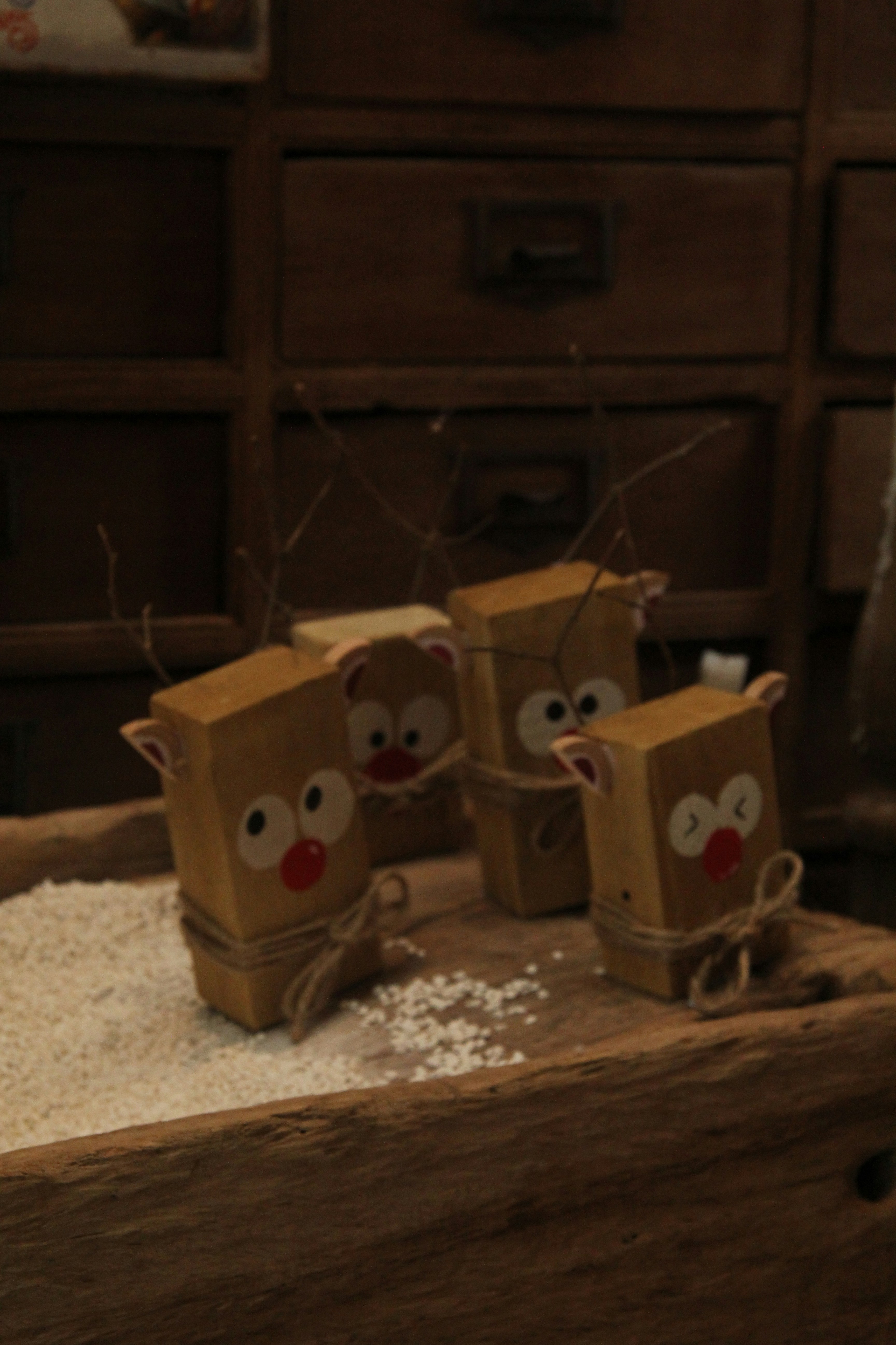 Wooden box reindeer toys with string antlers sit on a dusty wooden surface; a rustic cabinet backdrop and scattered white grains set a cozy, handmade scene.